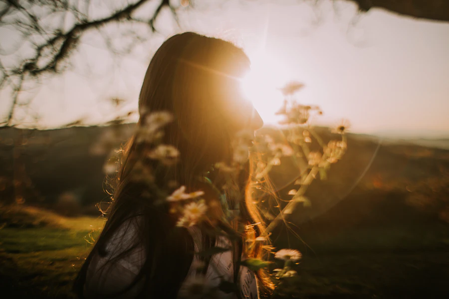 A woman in profile stands among blurred wildflowers with the sun flaring behind her head at sunset. Warm backlight outlines her hair against a soft hillside landscape.