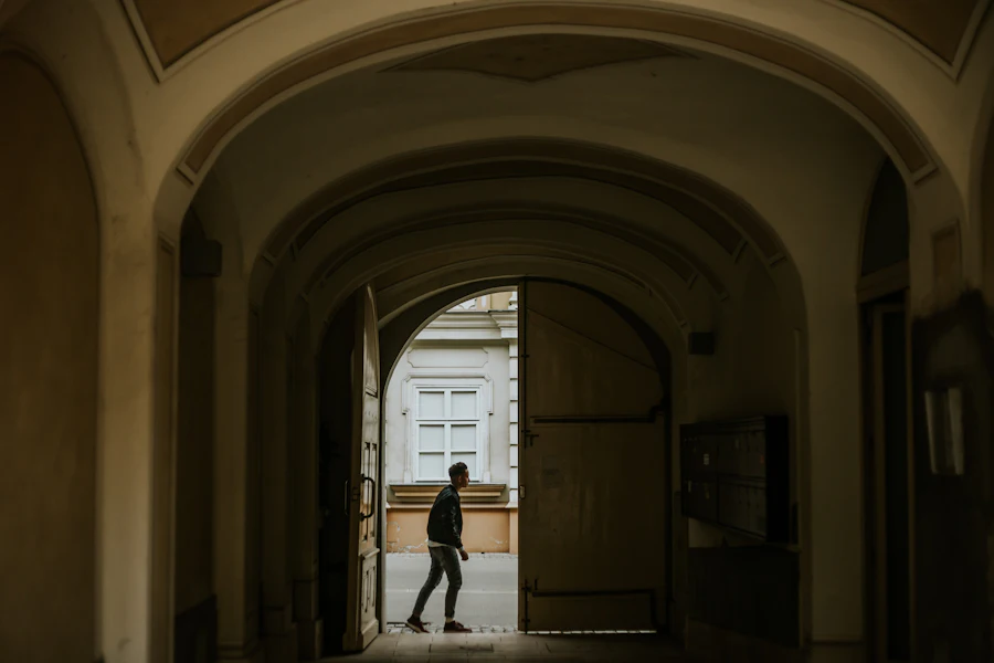 A person walks through a dark arched corridor toward a bright courtyard, framed by layered doorways and windows. The contrast emphasizes the silhouetted figure against the light outside.