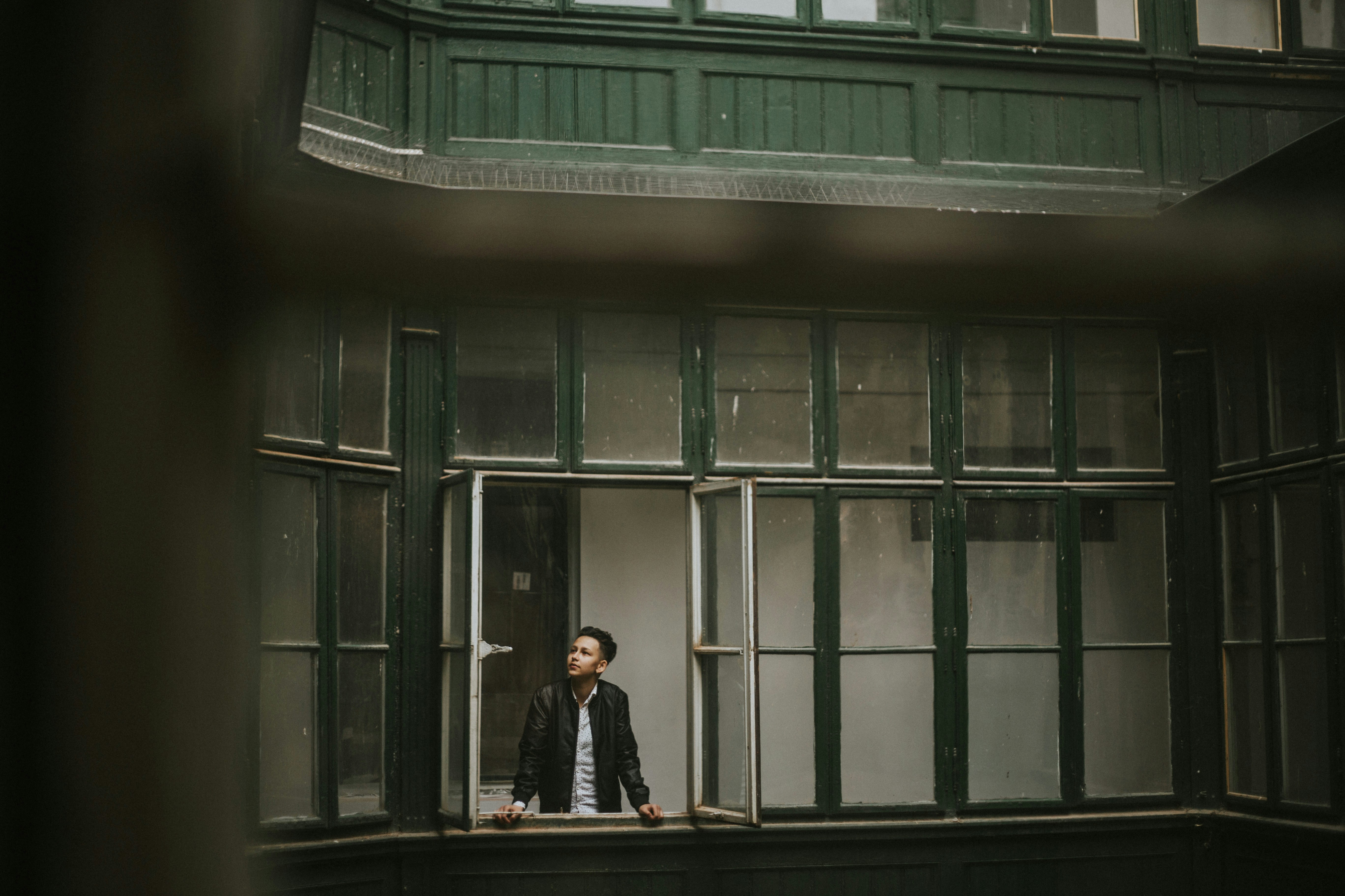 Man standing in a window of a vintage building, surrounded by green wooden frames, evoking a sense of quiet contemplation.