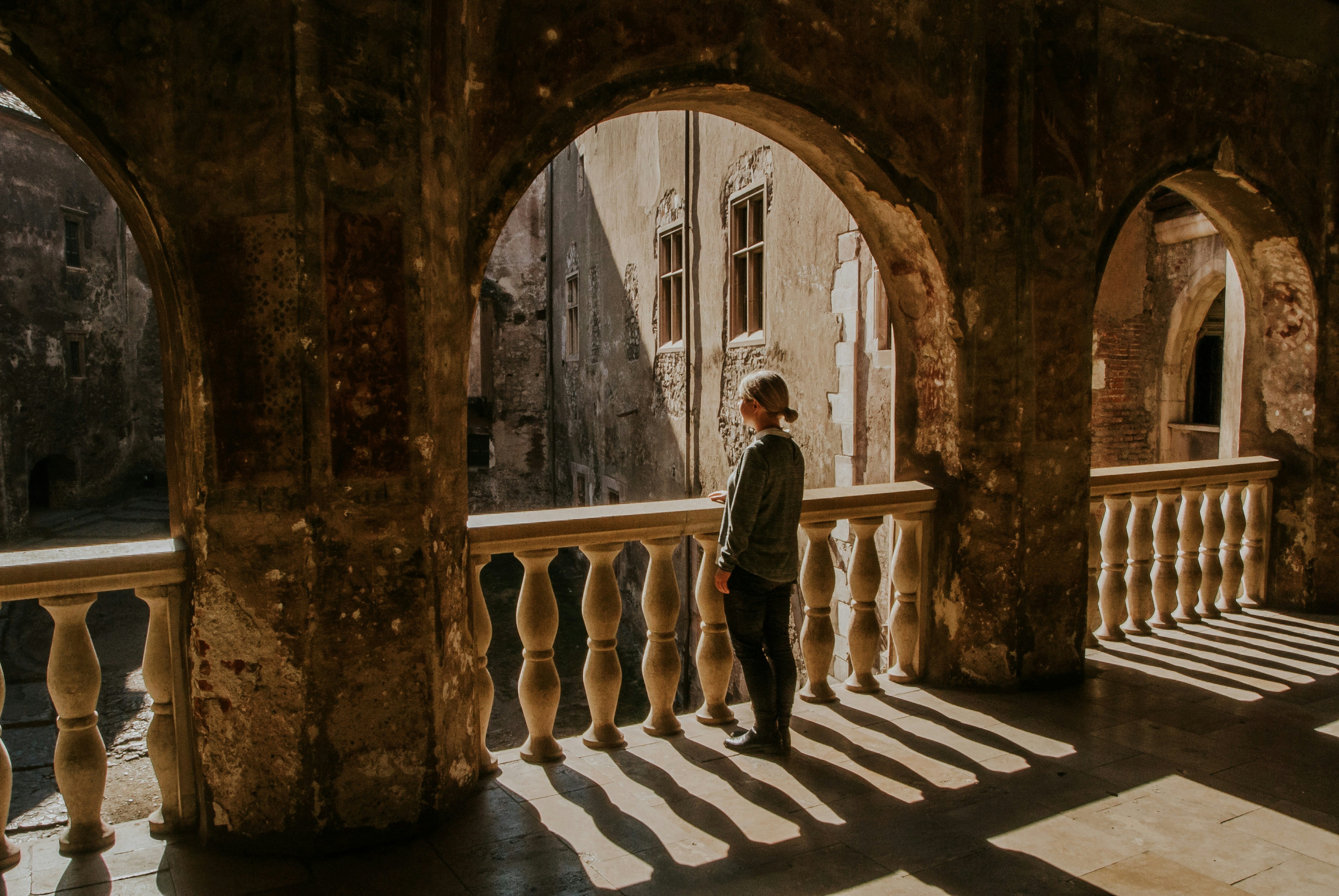 A figure stands pensively by a stone balustrade, framed by arched openings revealing a weathered courtyard. Shadows stretch across the floor, hinting at the passage of time.