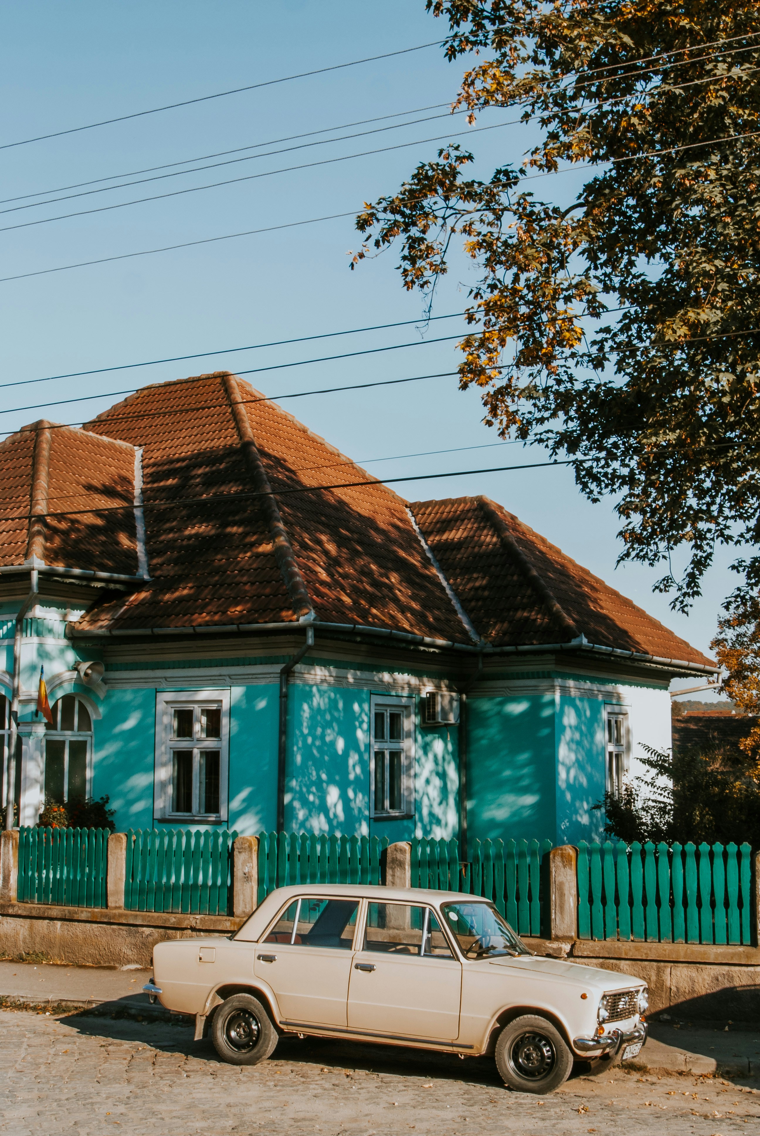 Photo of a vintage beige sedan parked on a cobbled street in front of a turquoise cottage with a red-tiled roof and teal fence.