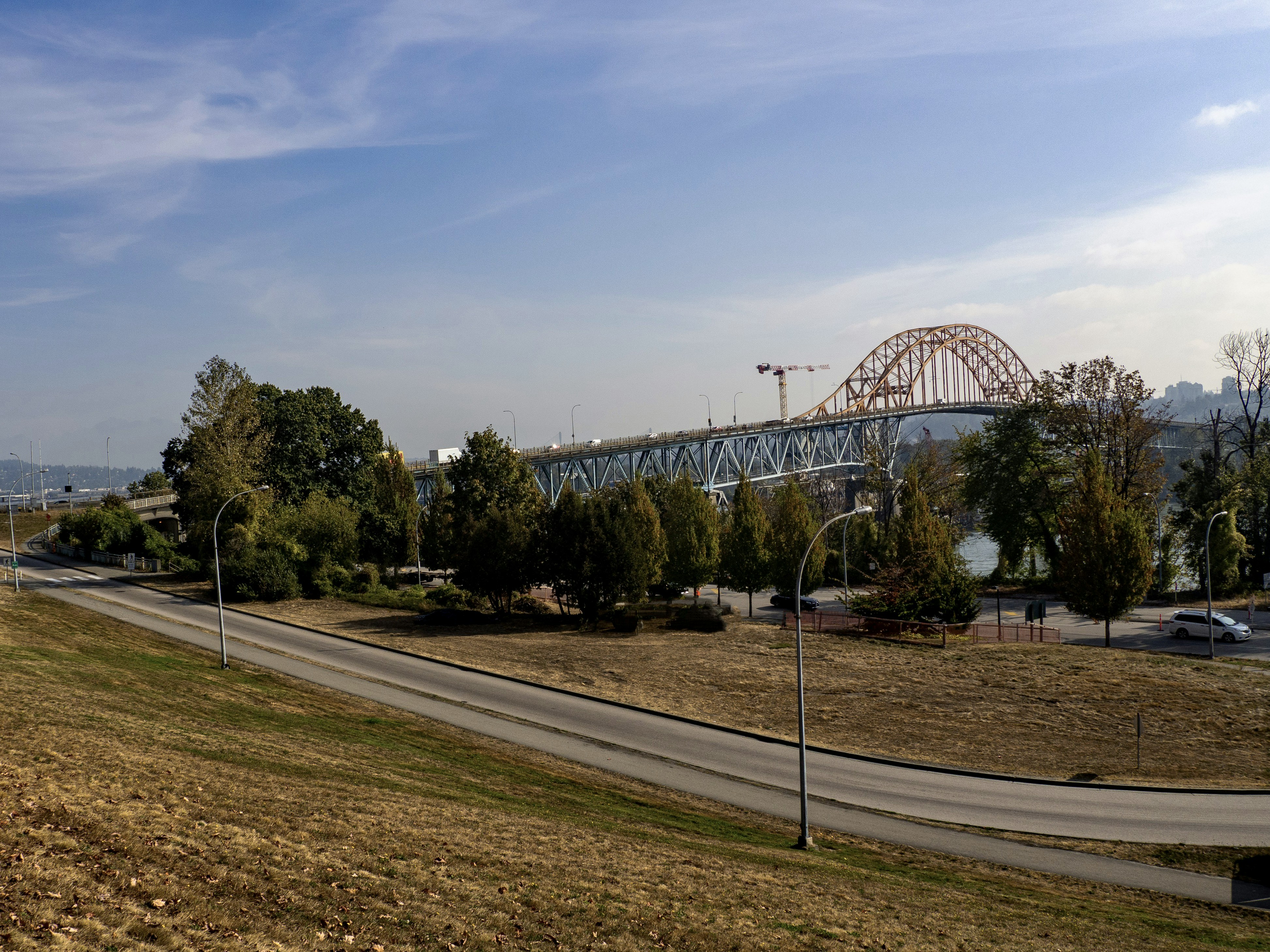 A scenic view of a bridge spanning a river, framed by trees and a gentle slope leading to a road. The scene captures the blend of natural and urban elements.