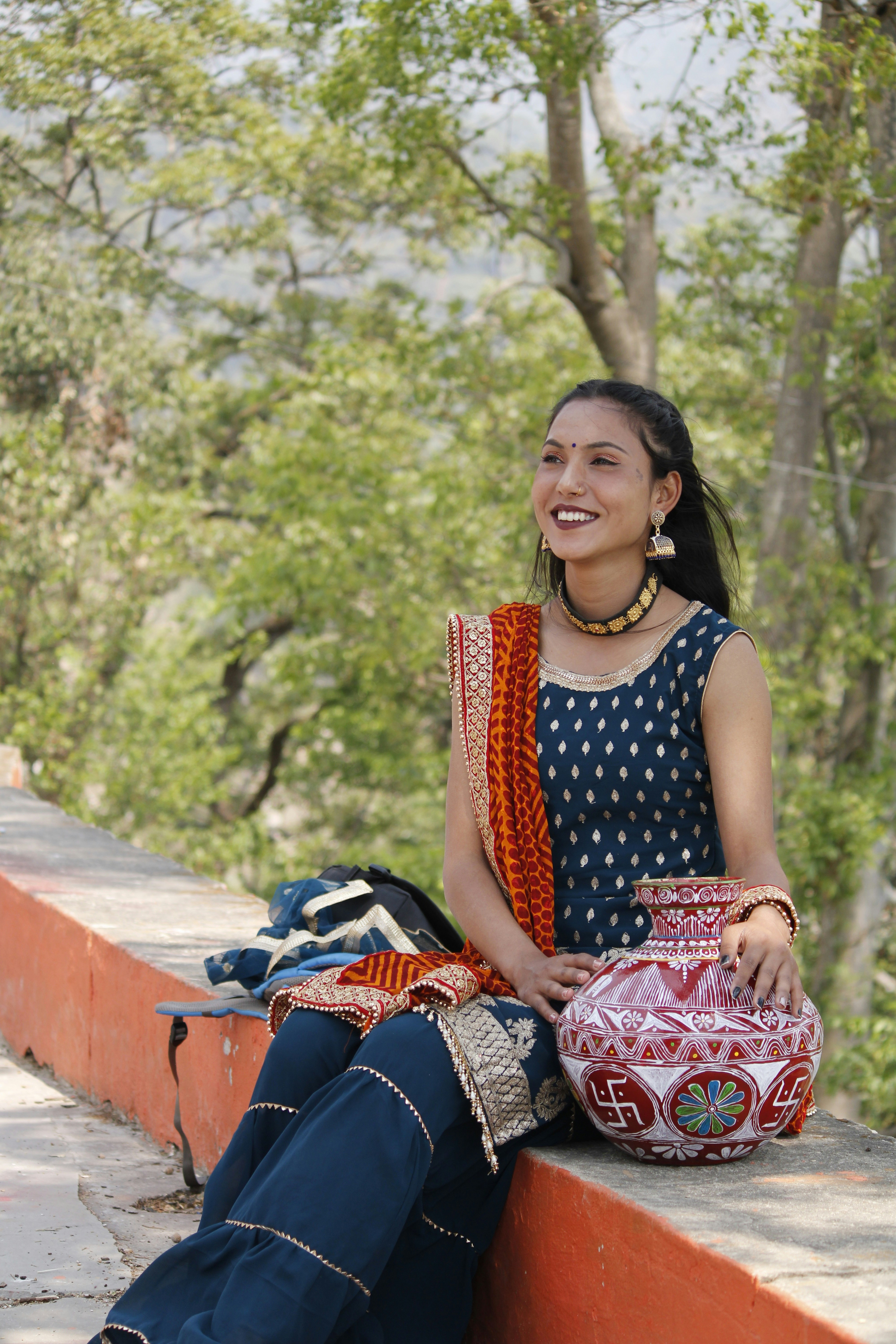 Woman in traditional attire sitting on a vibrant wall with a painted pot, surrounded by lush greenery.