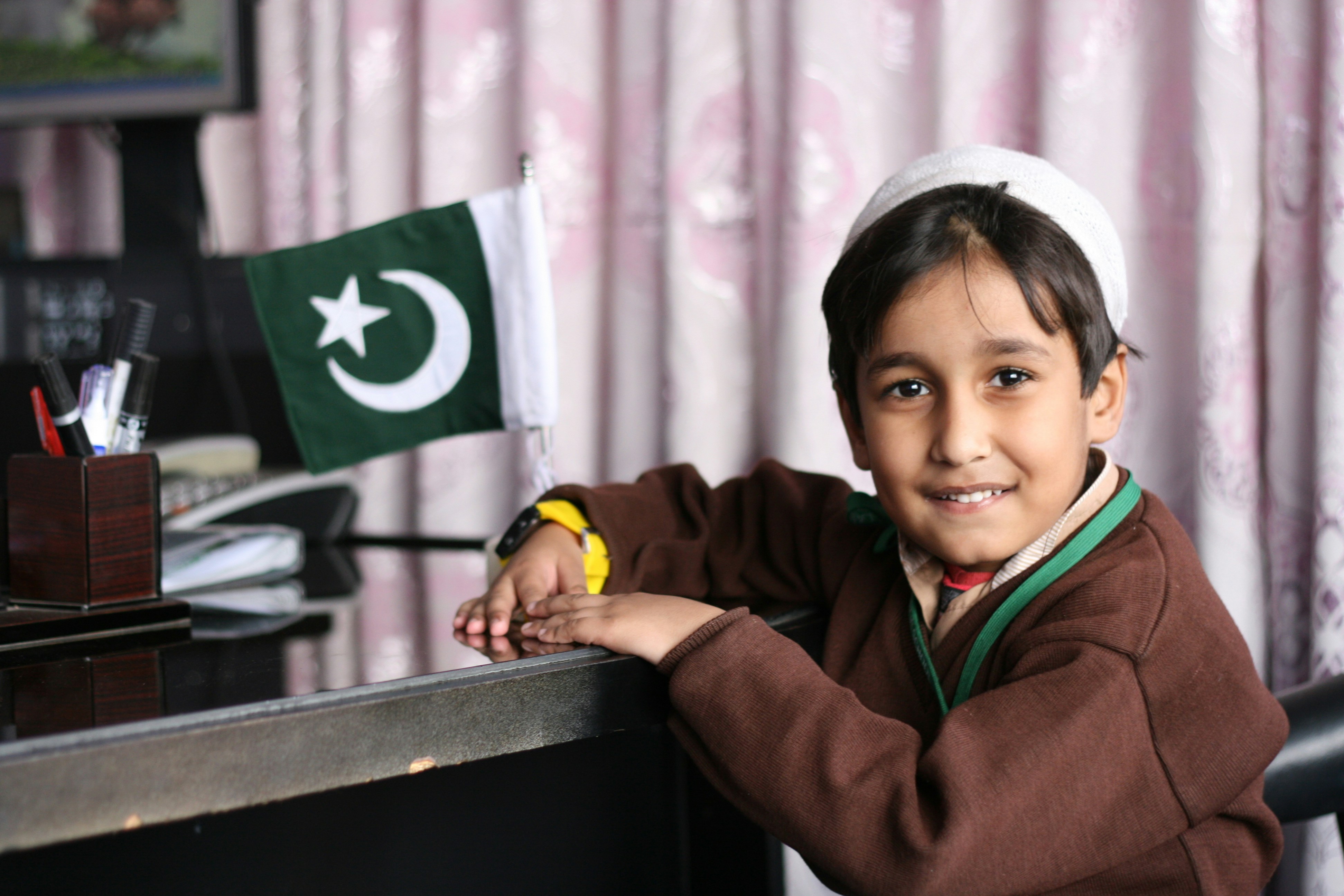 Smiling girl in a brown outfit holds a yellow flag beside a desk with a Pakistani flag.