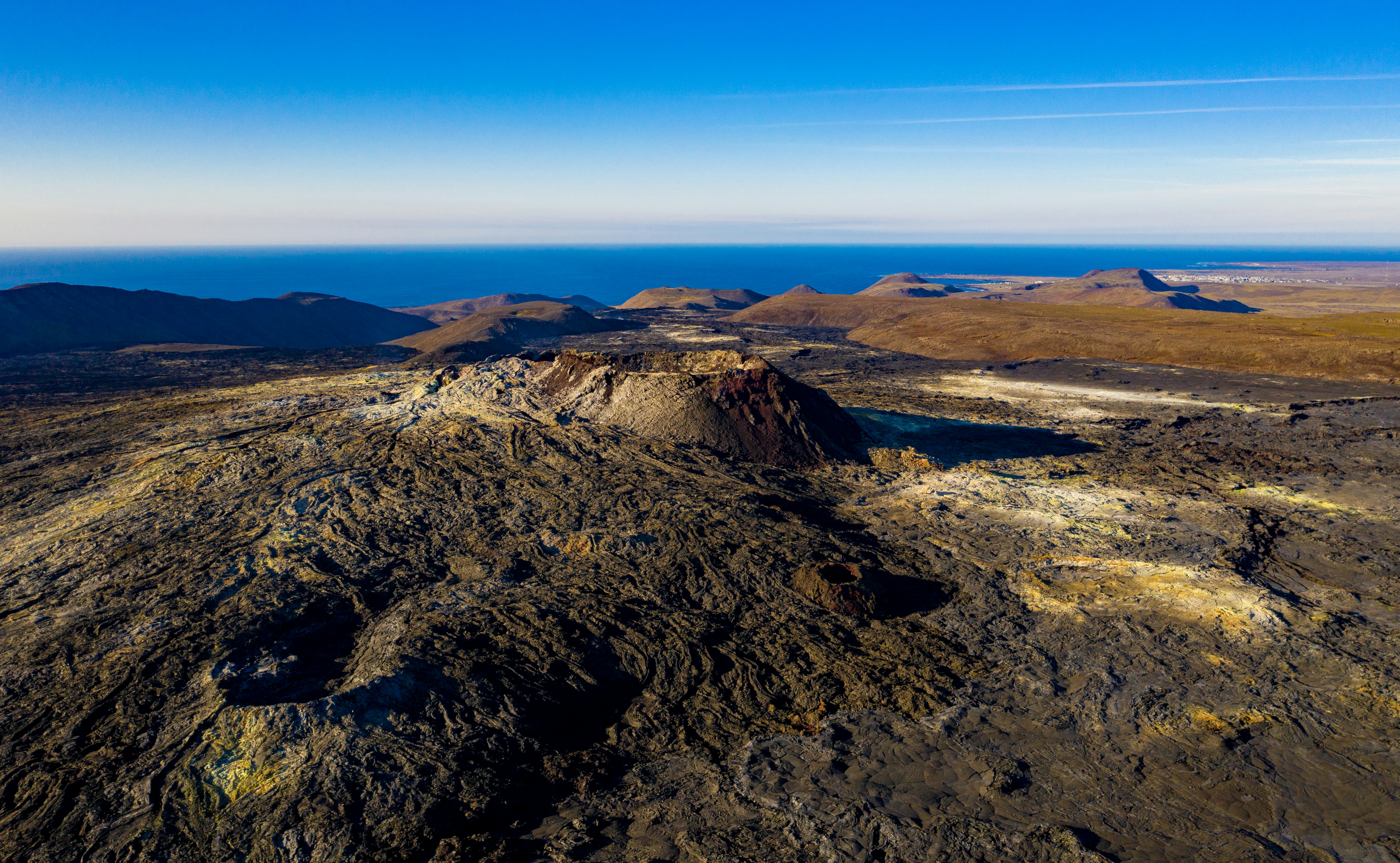 Aerial view of a volcanic landscape showcasing a rugged terrain with a central crater and distant ocean views. The stark contrast between the dark lava and the serene blue sea is striking.