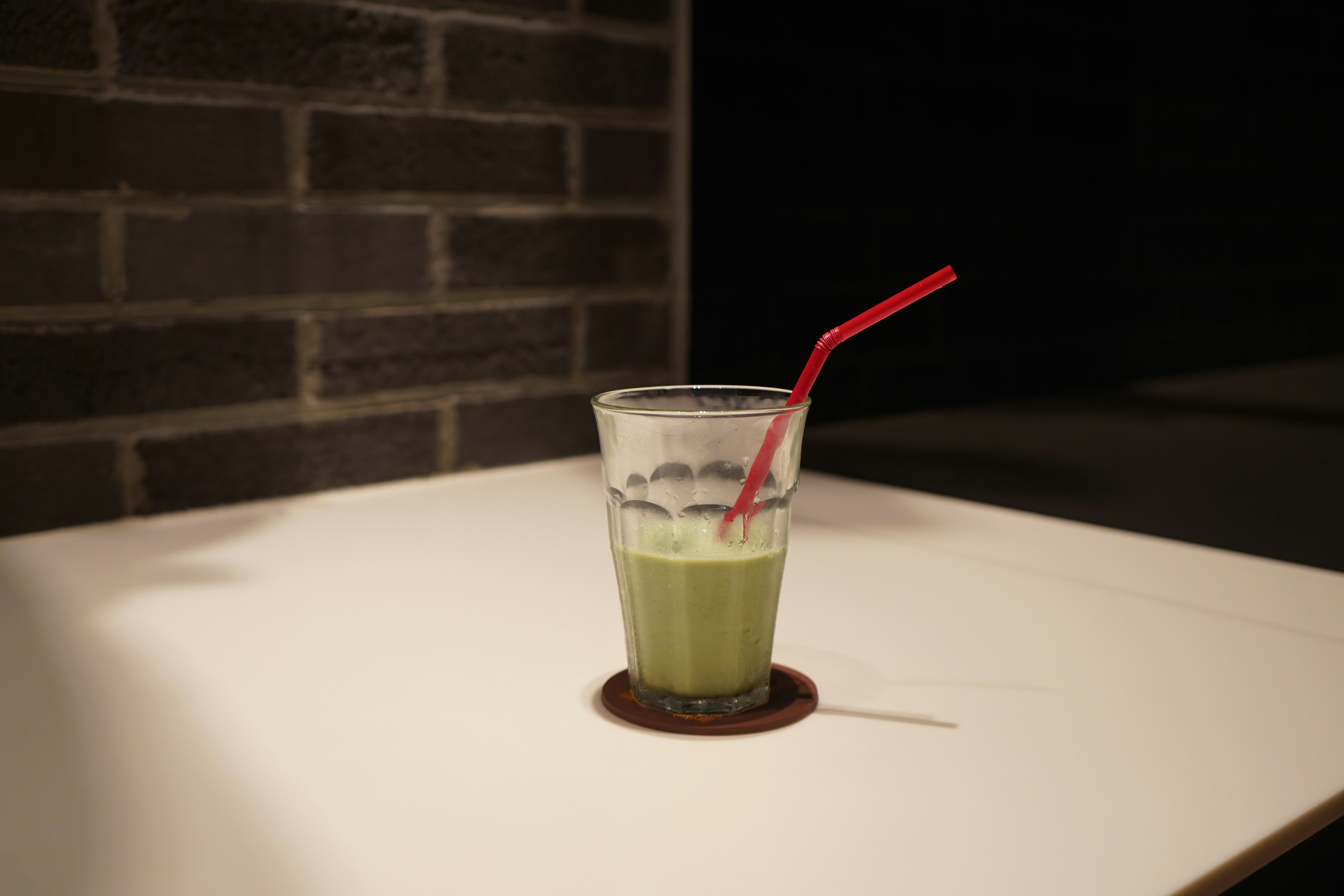 Glass of matcha with a red straw resting on a coaster atop a white table, surrounded by a textured brick wall.
