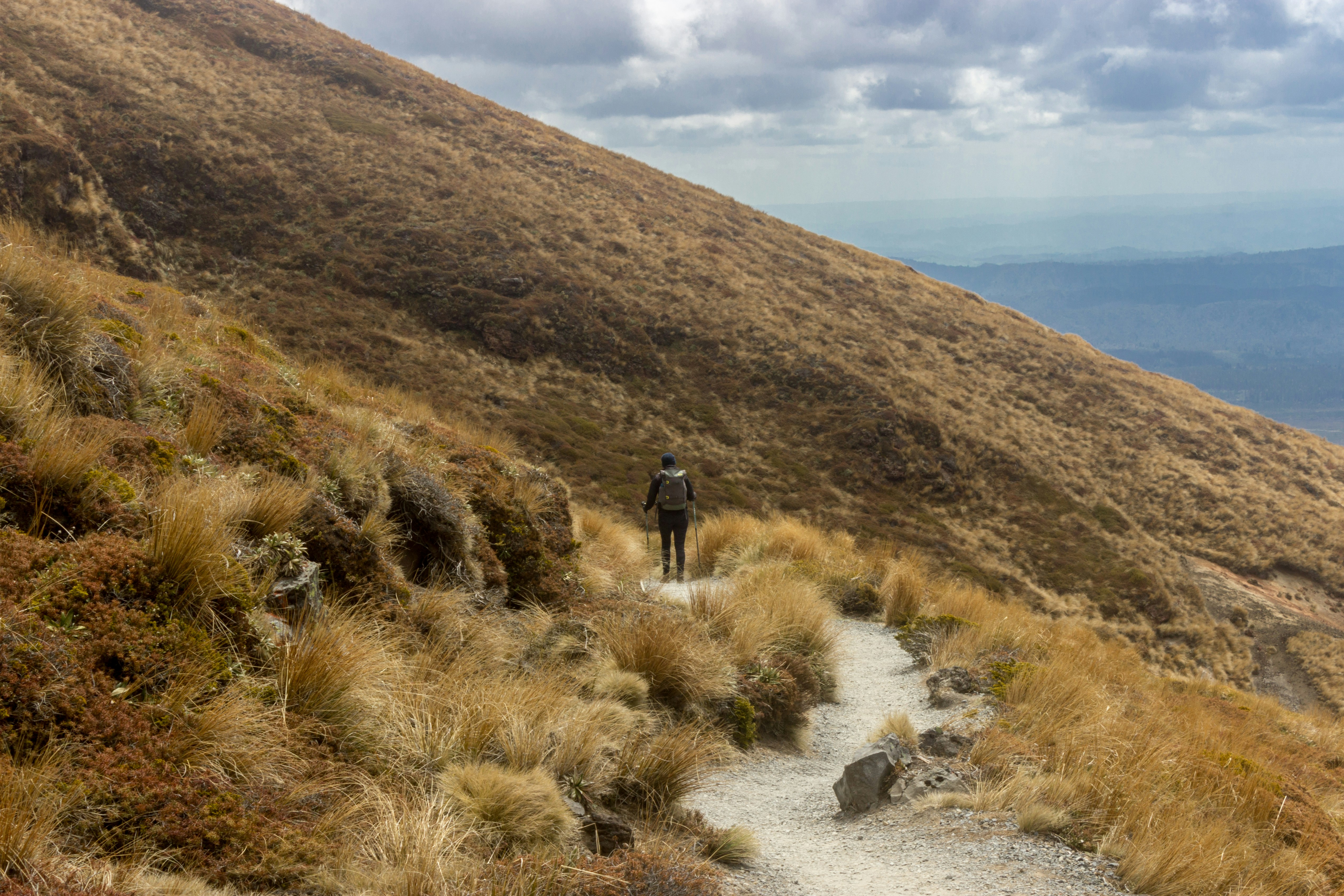 Hiker on a narrow trail surrounded by rocky terrain and golden grasses under a cloudy sky.