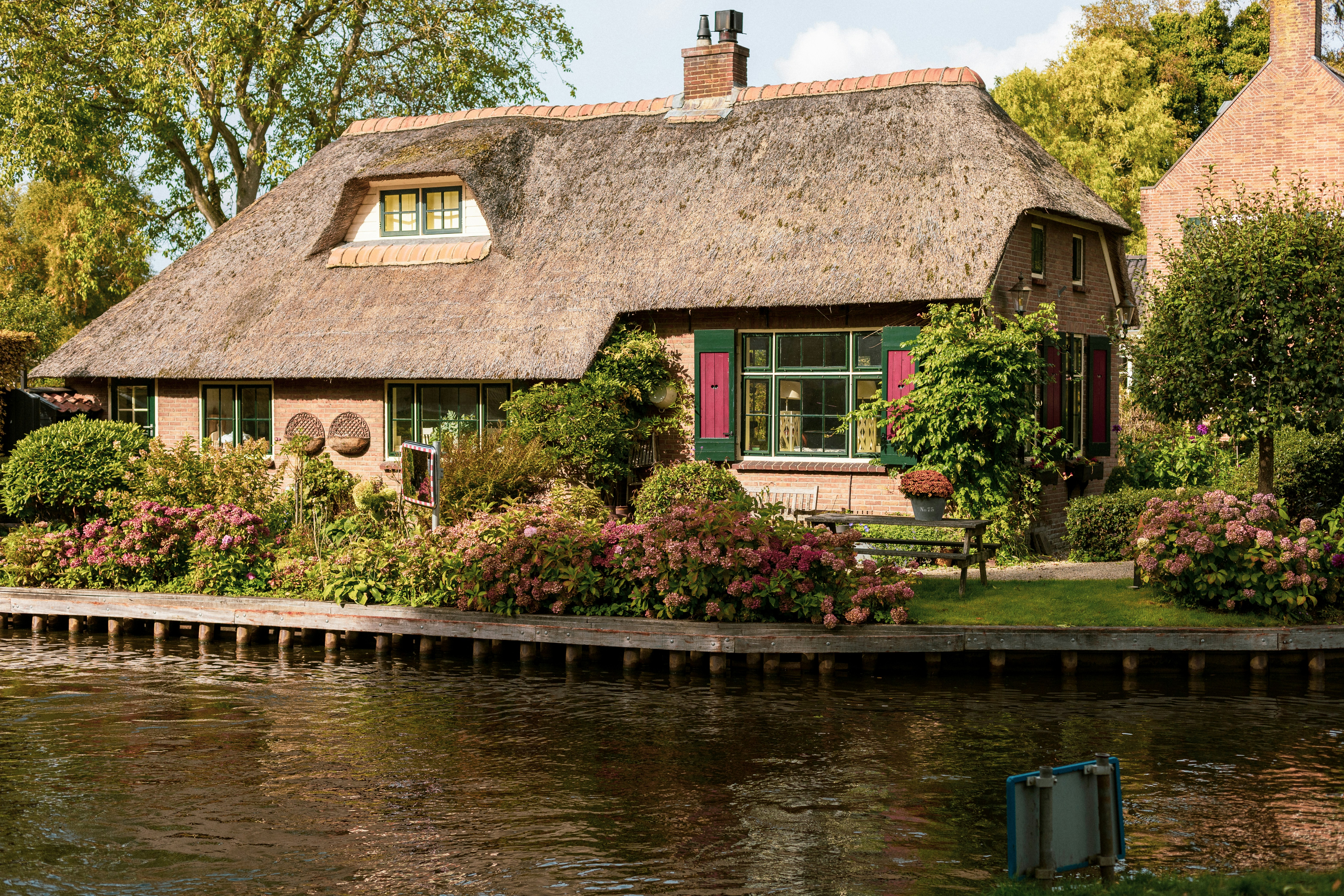 Quaint thatched cottage surrounded by lush greenery and vibrant flowers beside a tranquil river.