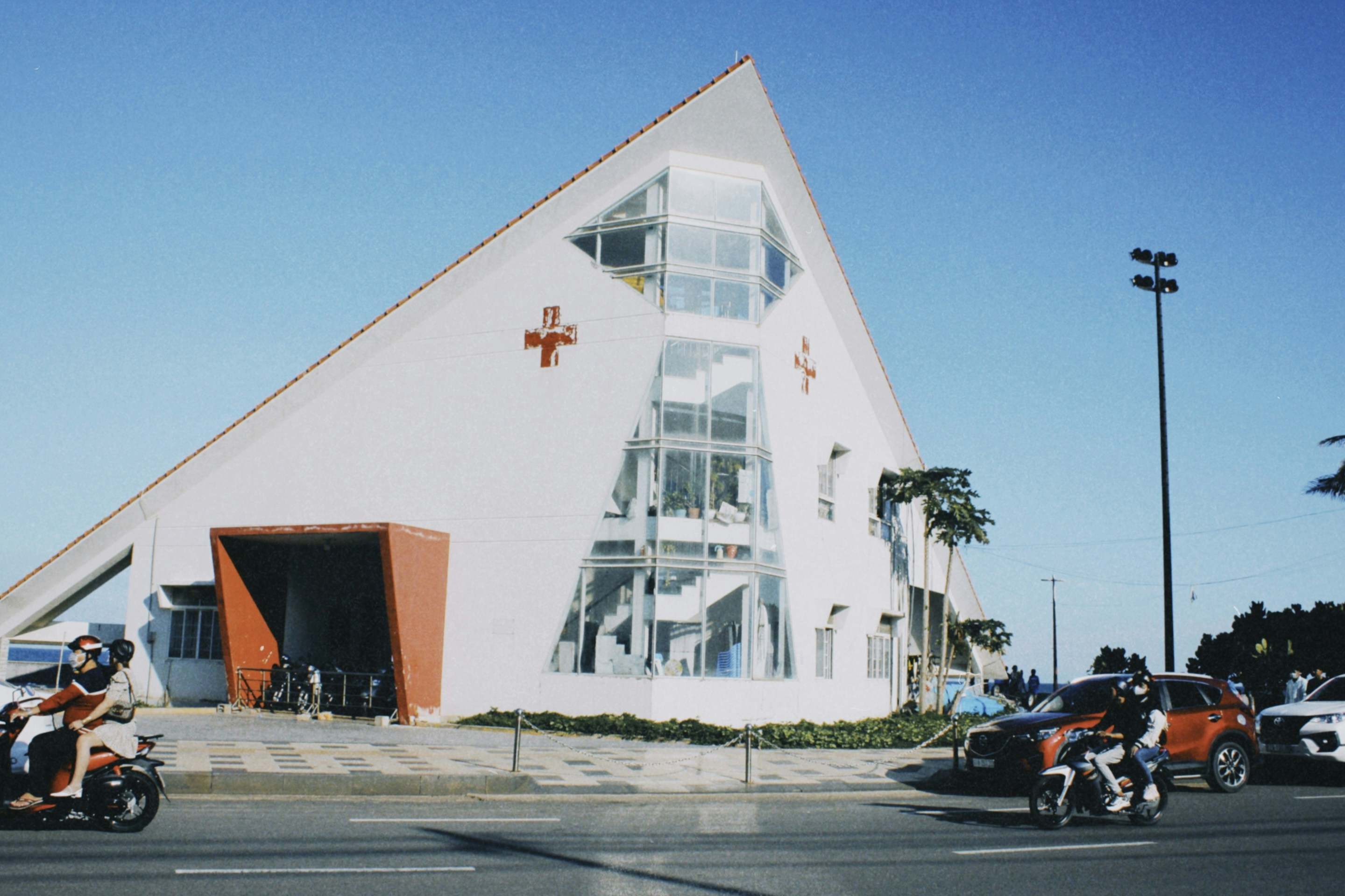 Contemporary building with a striking triangular design, featuring large glass panels and vibrant red accents, situated along a bustling street.