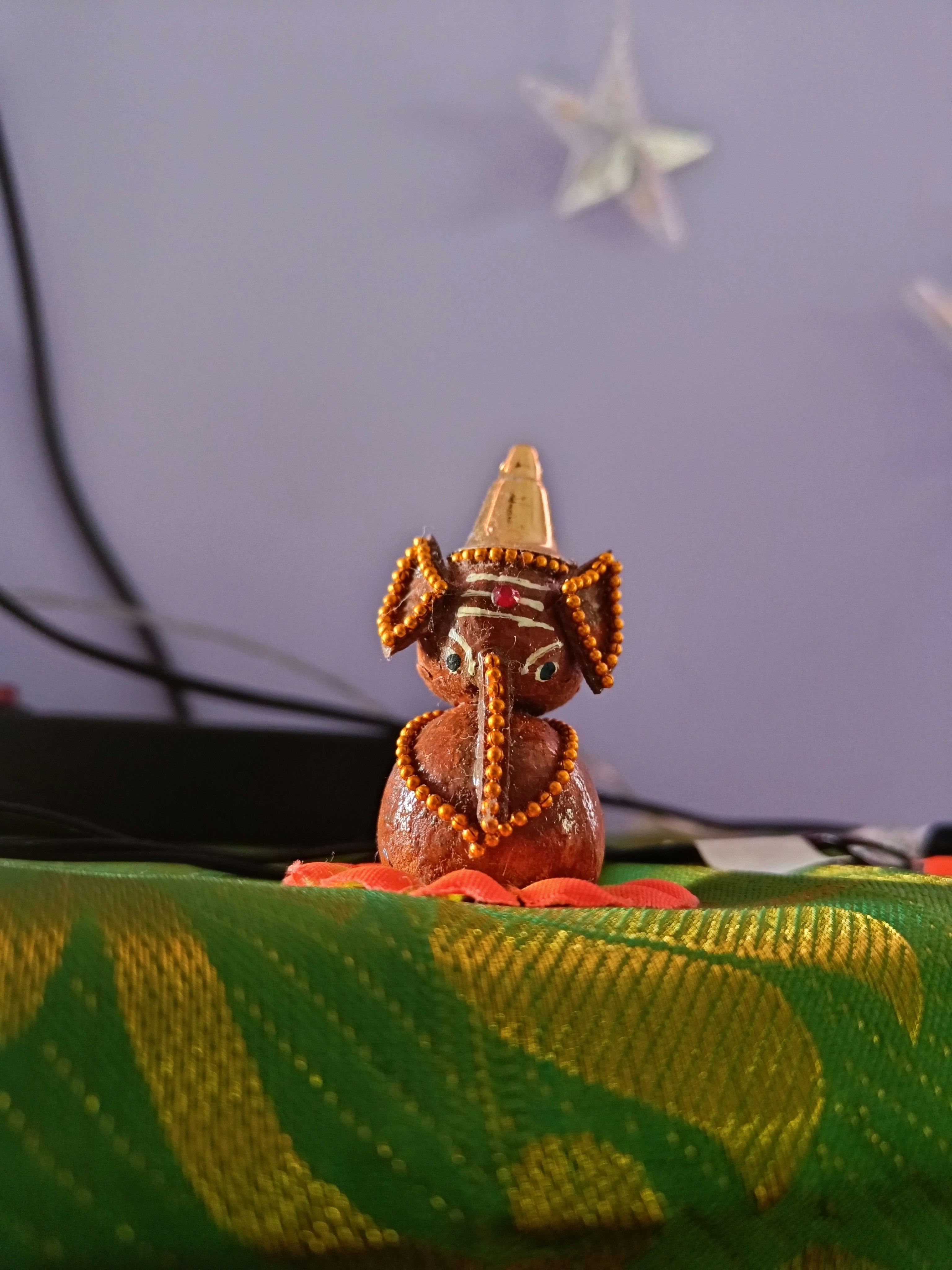 Handcrafted Ganesh idol adorned with intricate details, resting on vibrant fabric. Background features soft, blurred elements enhancing the focus on the idol.