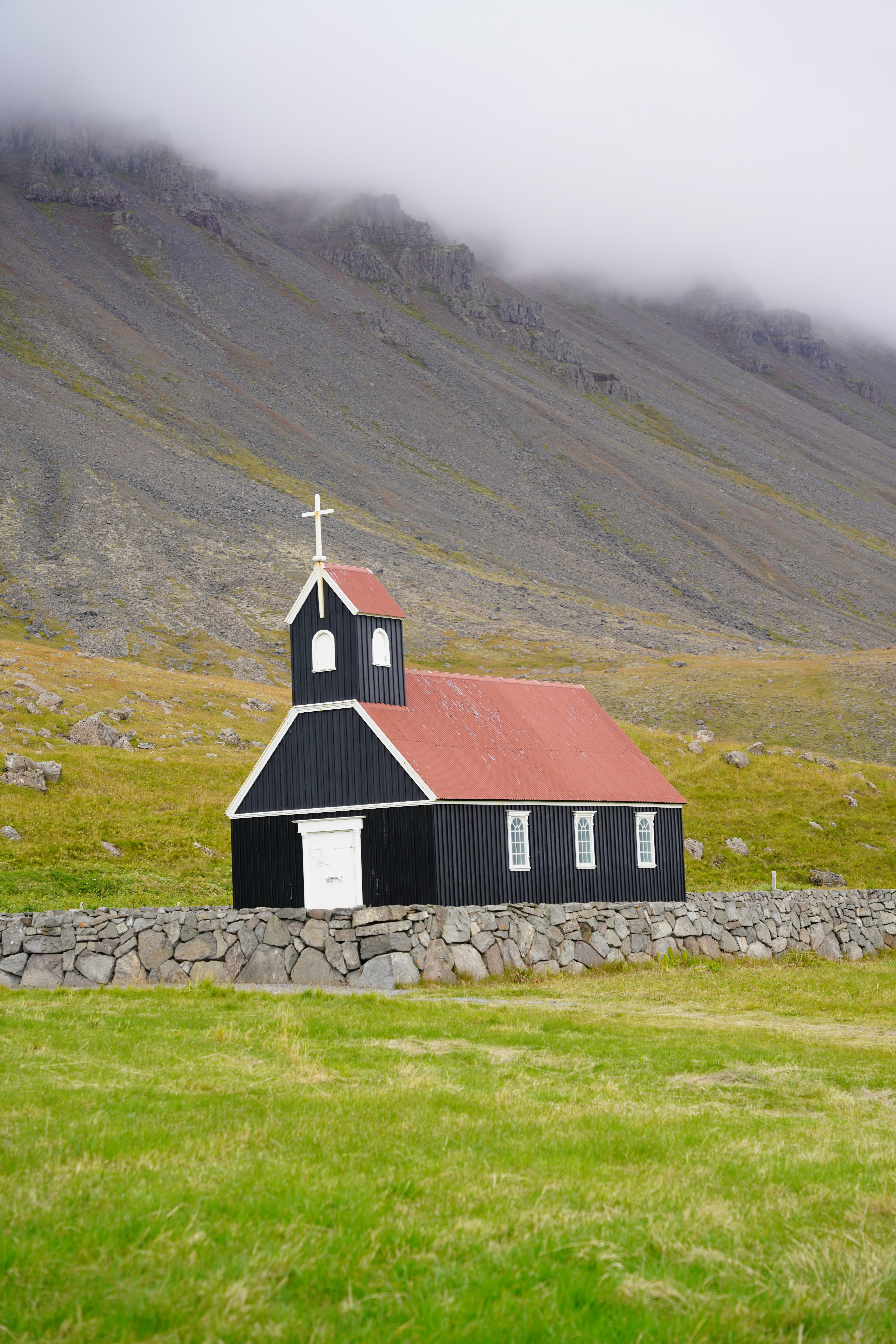 Black wooden church with a red roof stands against a backdrop of rocky mountains and mist. The surrounding green grass adds a touch of vibrancy.