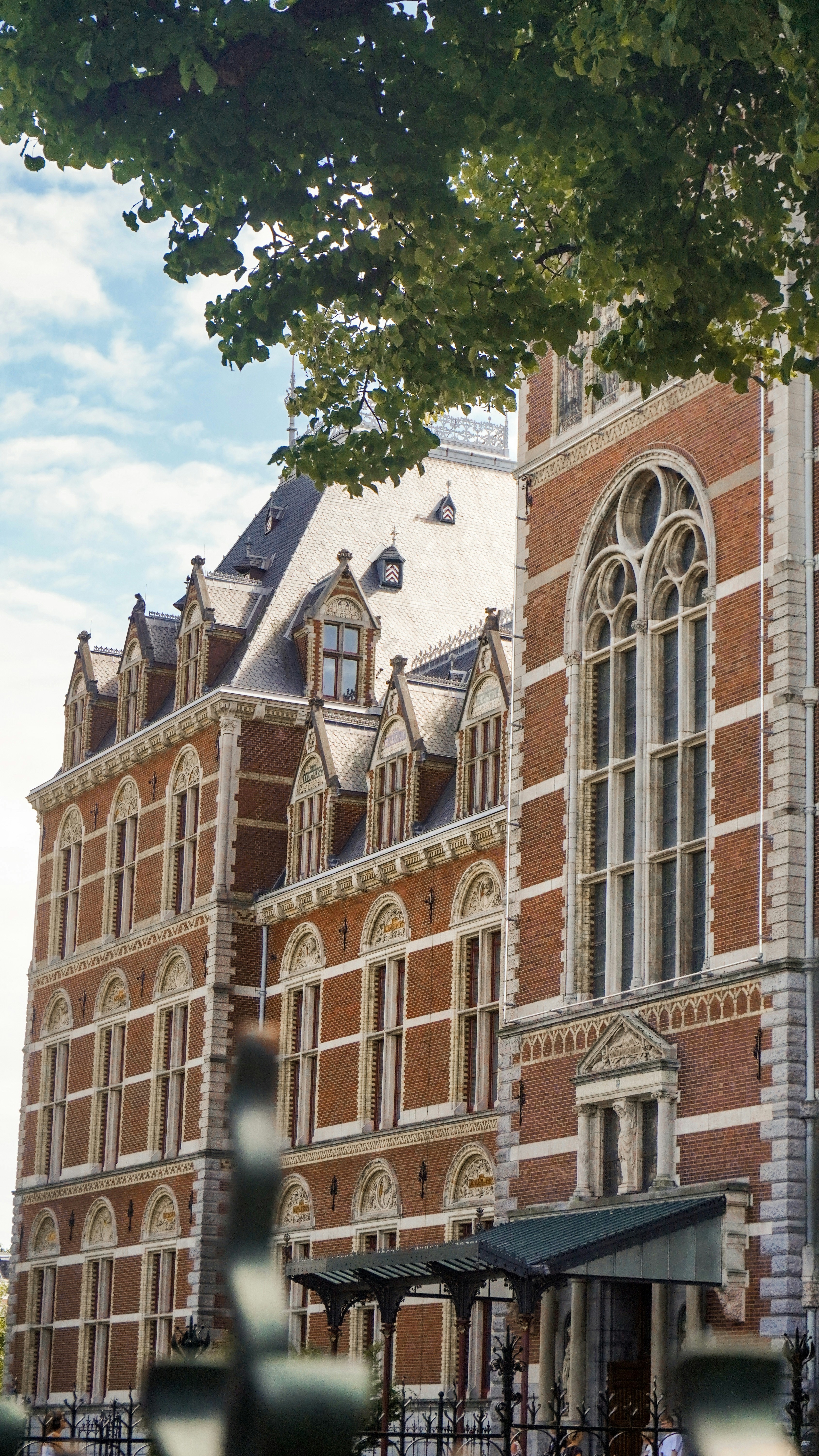 Historic brick building with ornate windows and detailed architecture, partially framed by lush green leaves. 