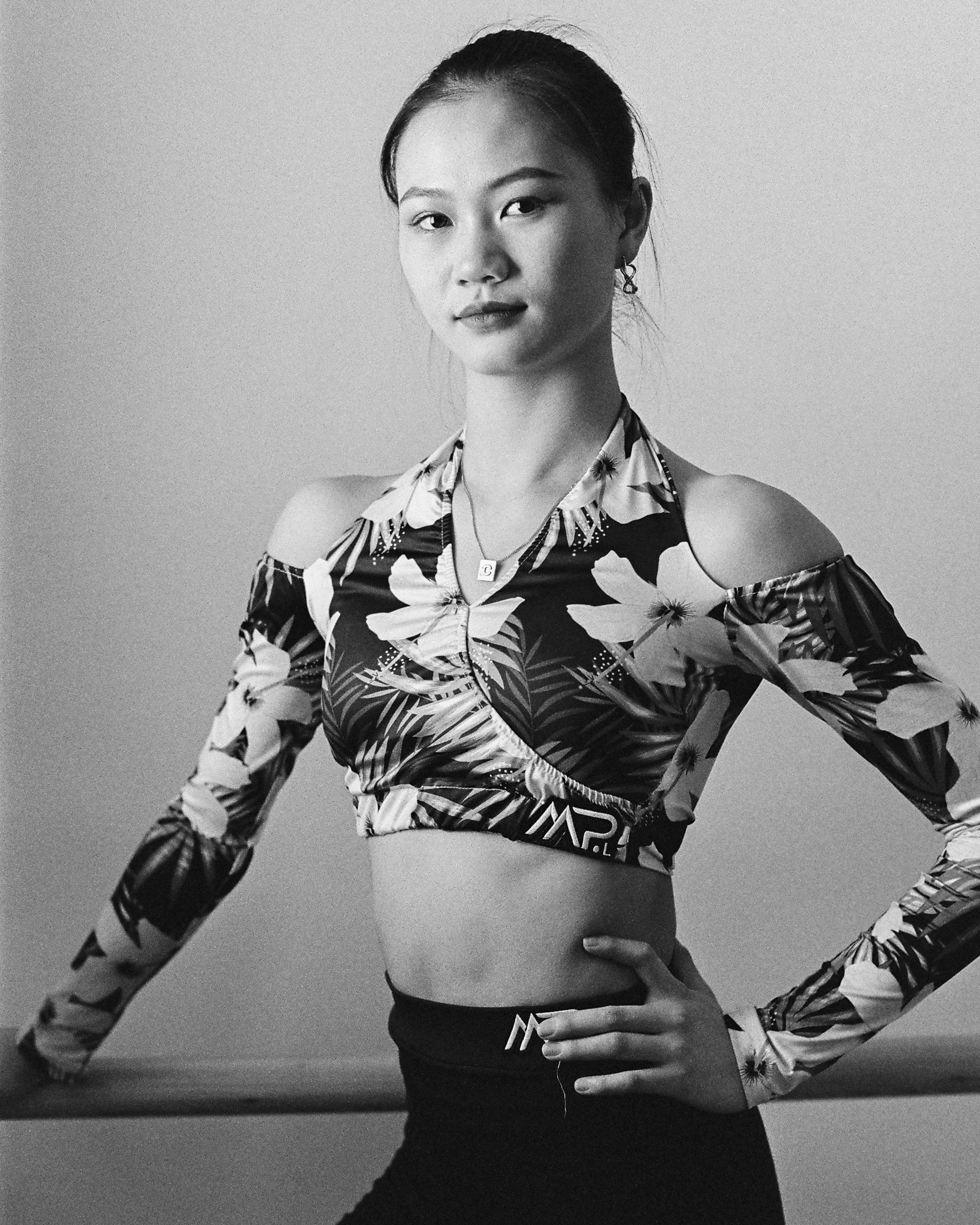 Black-and-white portrait of a dancer posing at a barre in a floral cropped top, with one hand on the barre and the other on the hip.