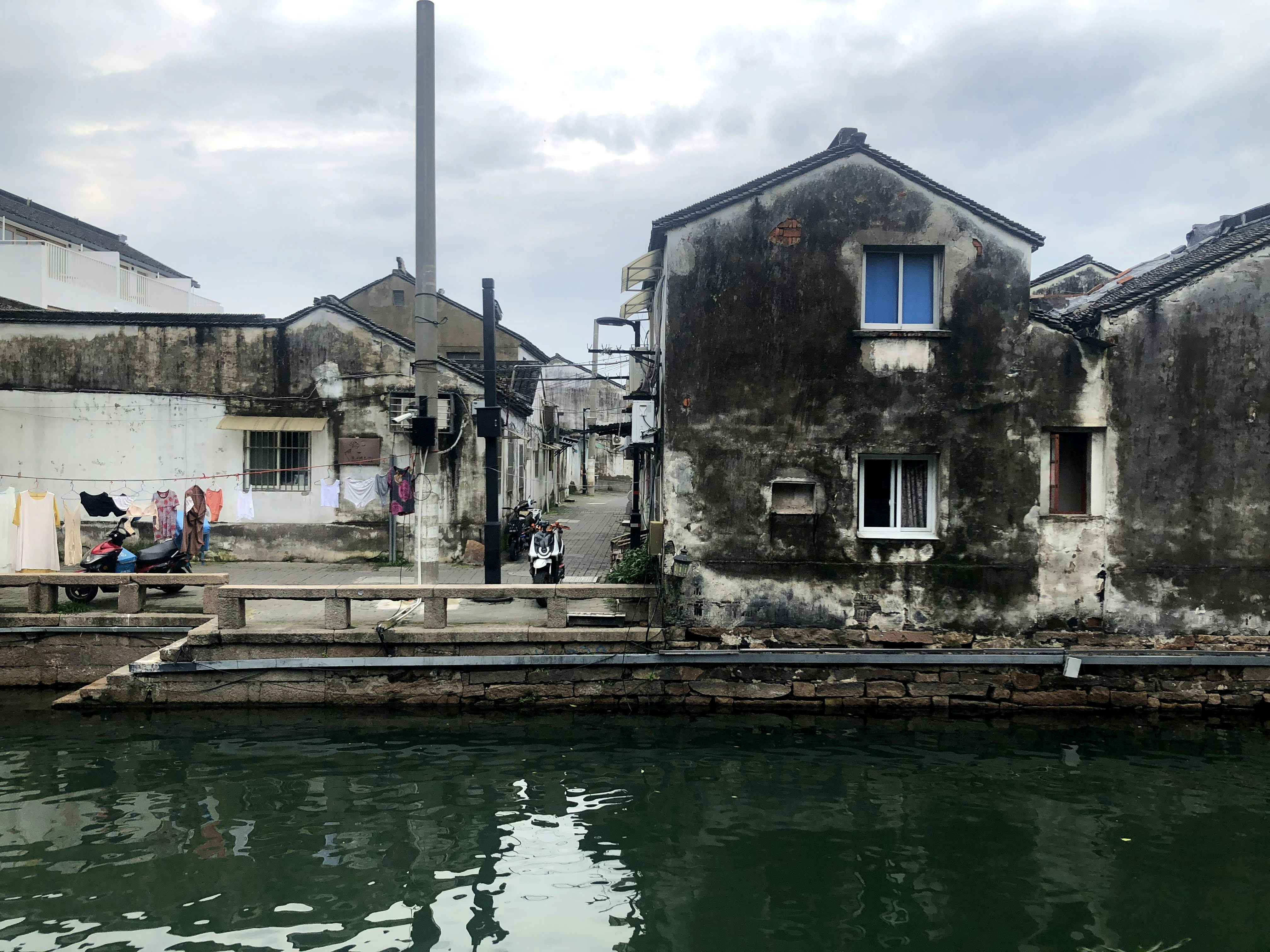Rustic buildings with weathered facades line a tranquil canal under a cloudy sky.