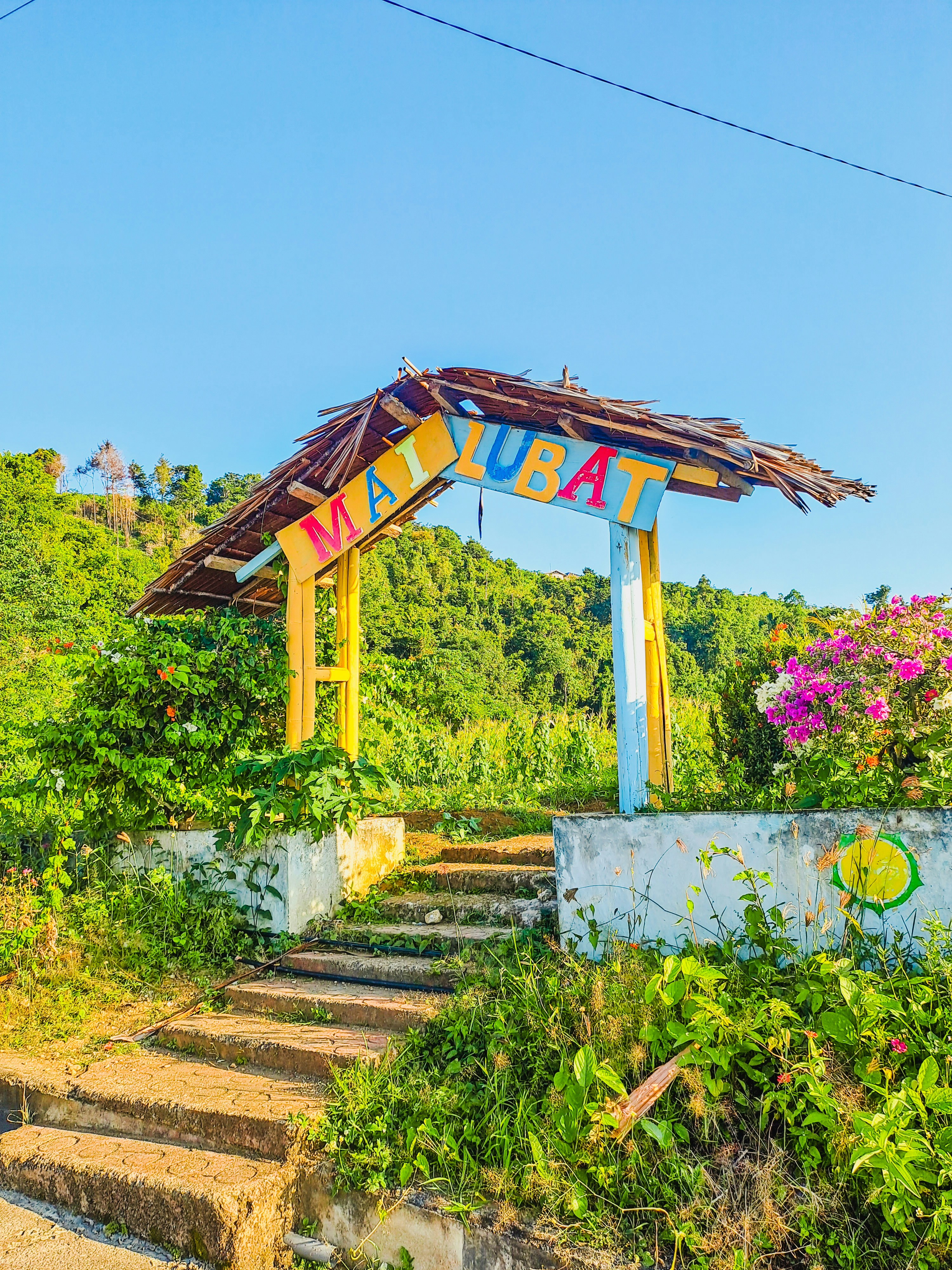 Vibrant archway sign reading 'MALLUBAT' framed by lush greenery and colorful flowers, inviting visitors to explore the area.