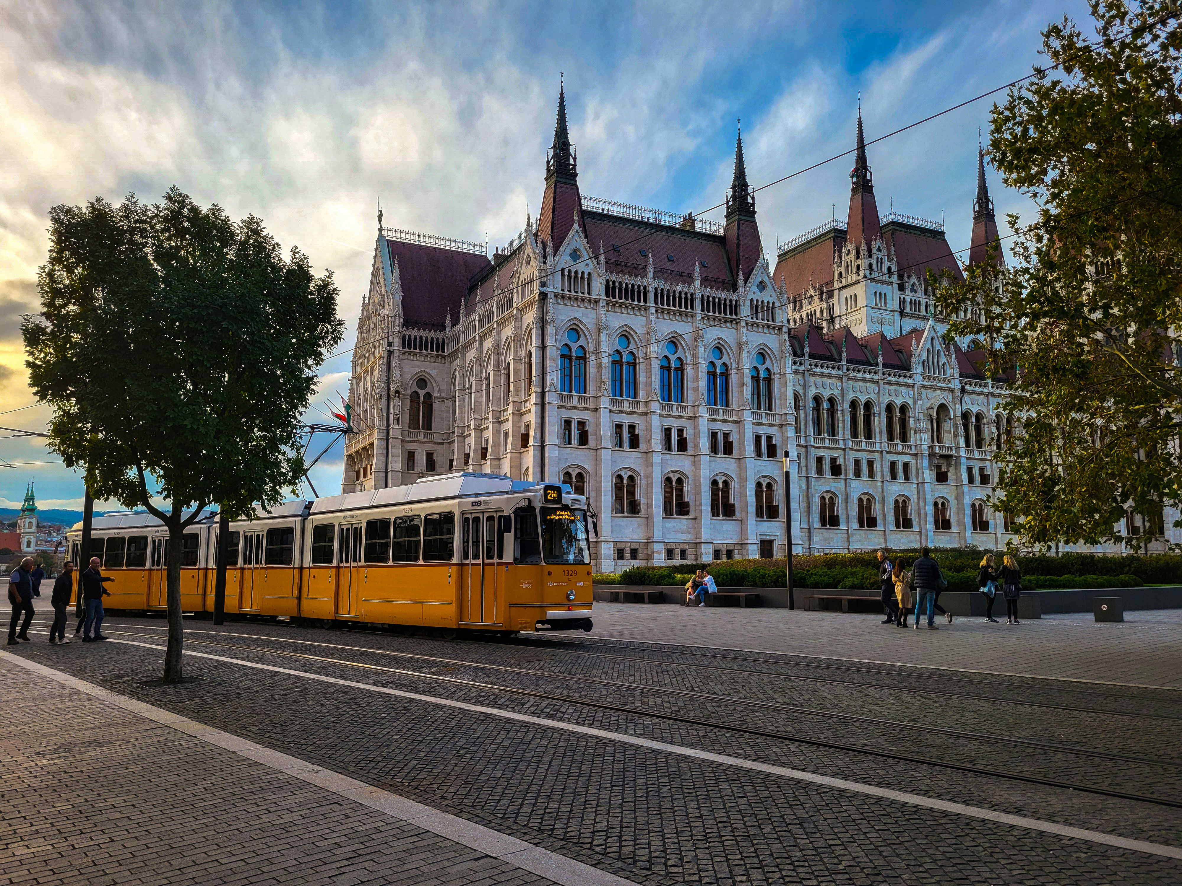 Yellow tram gliding past the ornate facade of the Hungarian Parliament Building under a dramatic sky.
