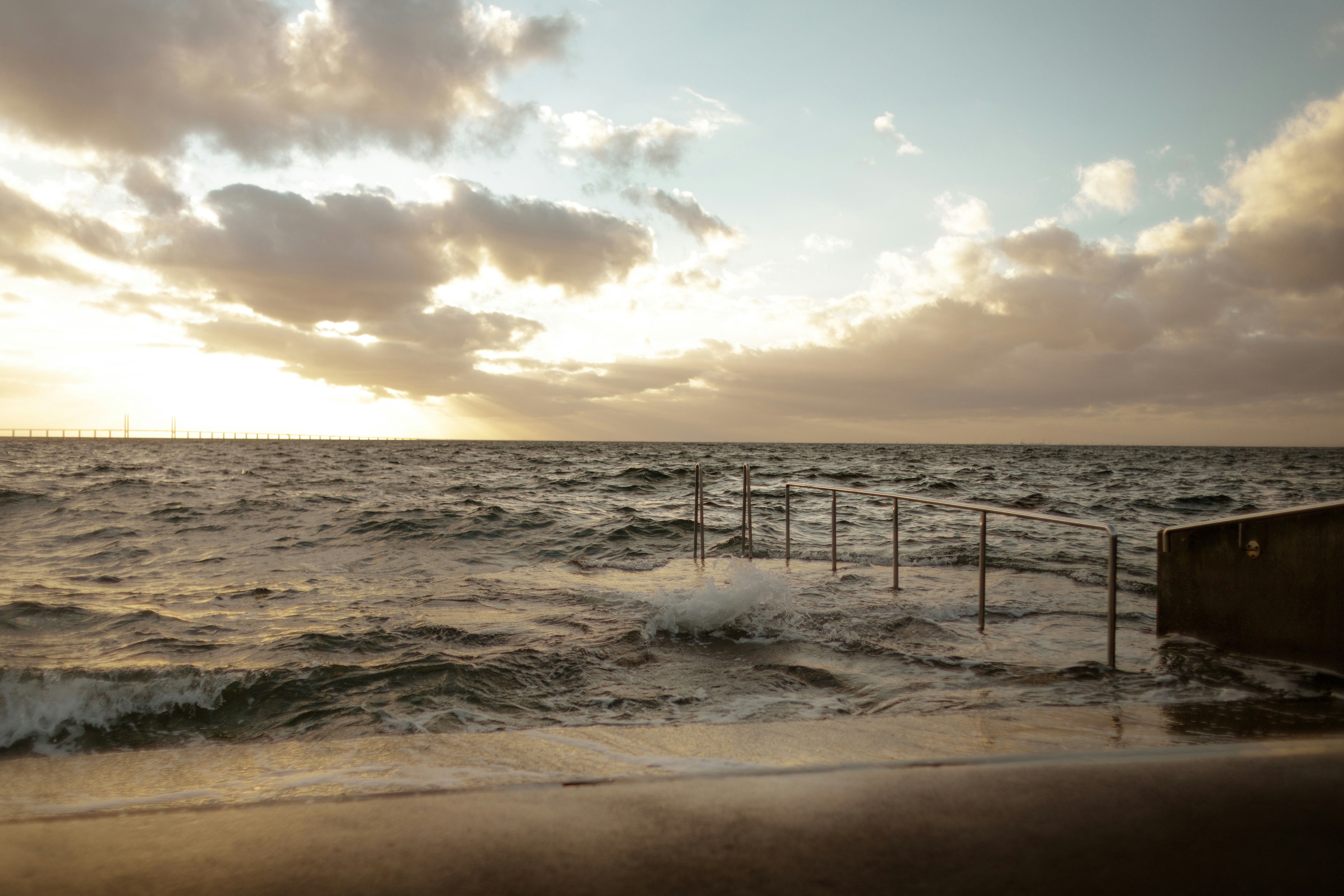 Ocean waves crash against a sandy shore under a dramatic sky at sunset.