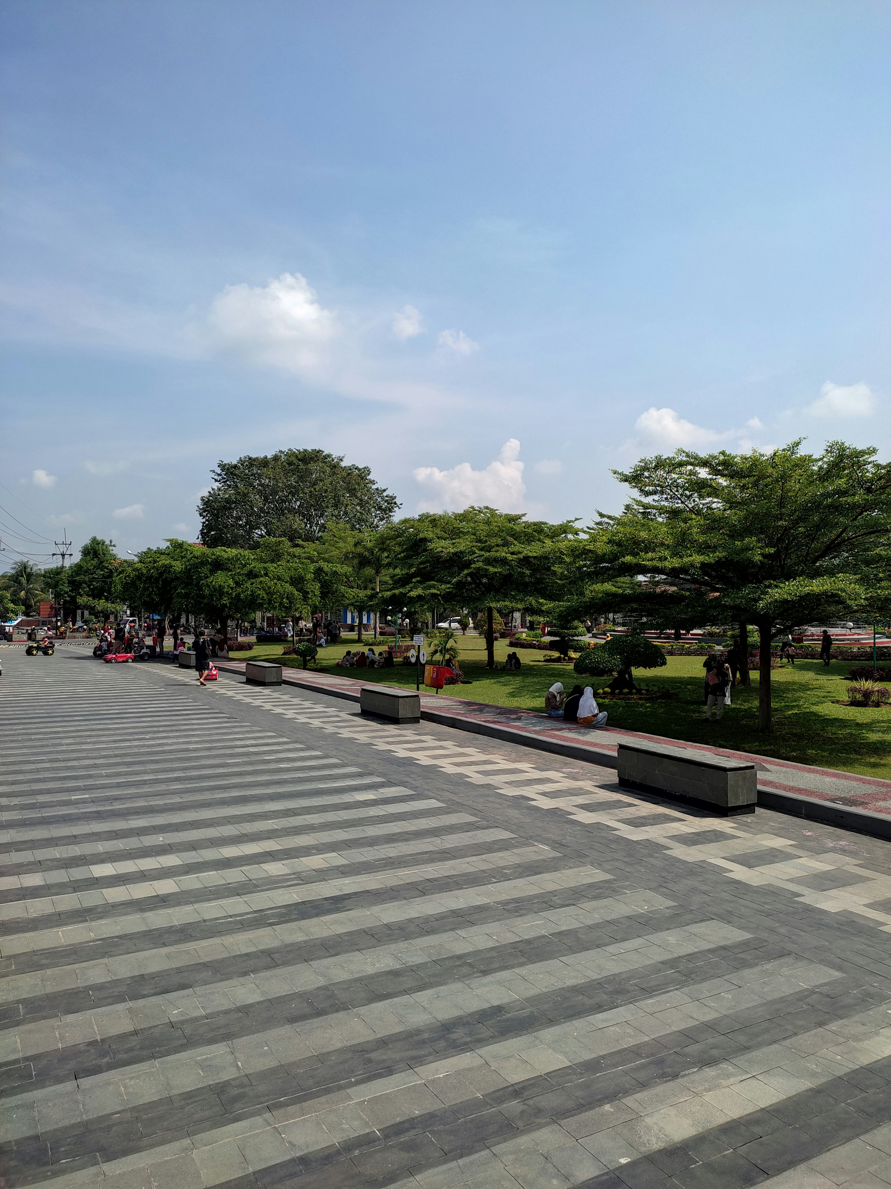 People enjoying leisure time in a park filled with lush greenery and patterned pathways. The scene captures a vibrant community atmosphere.