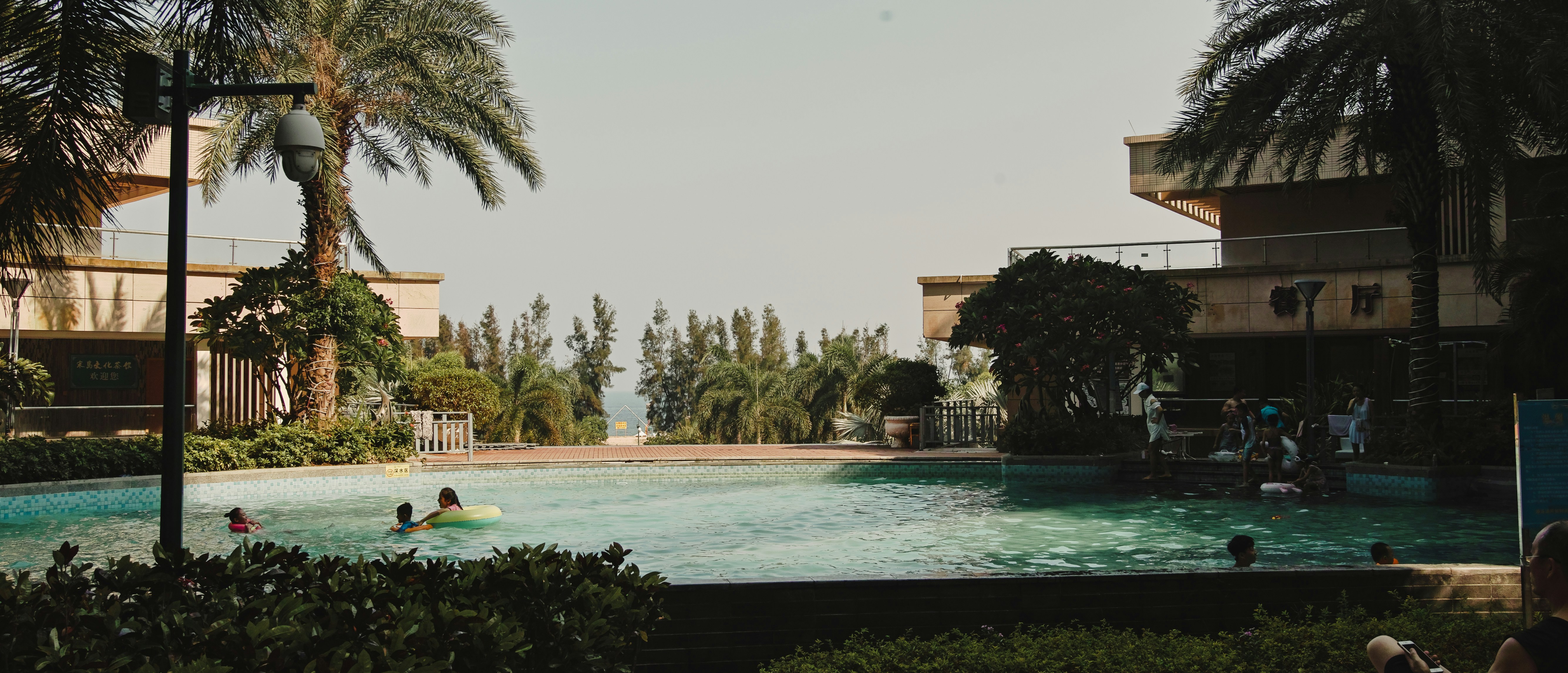 Palm-fringed pool with swimmers, framed by resort buildings under a clear sky.