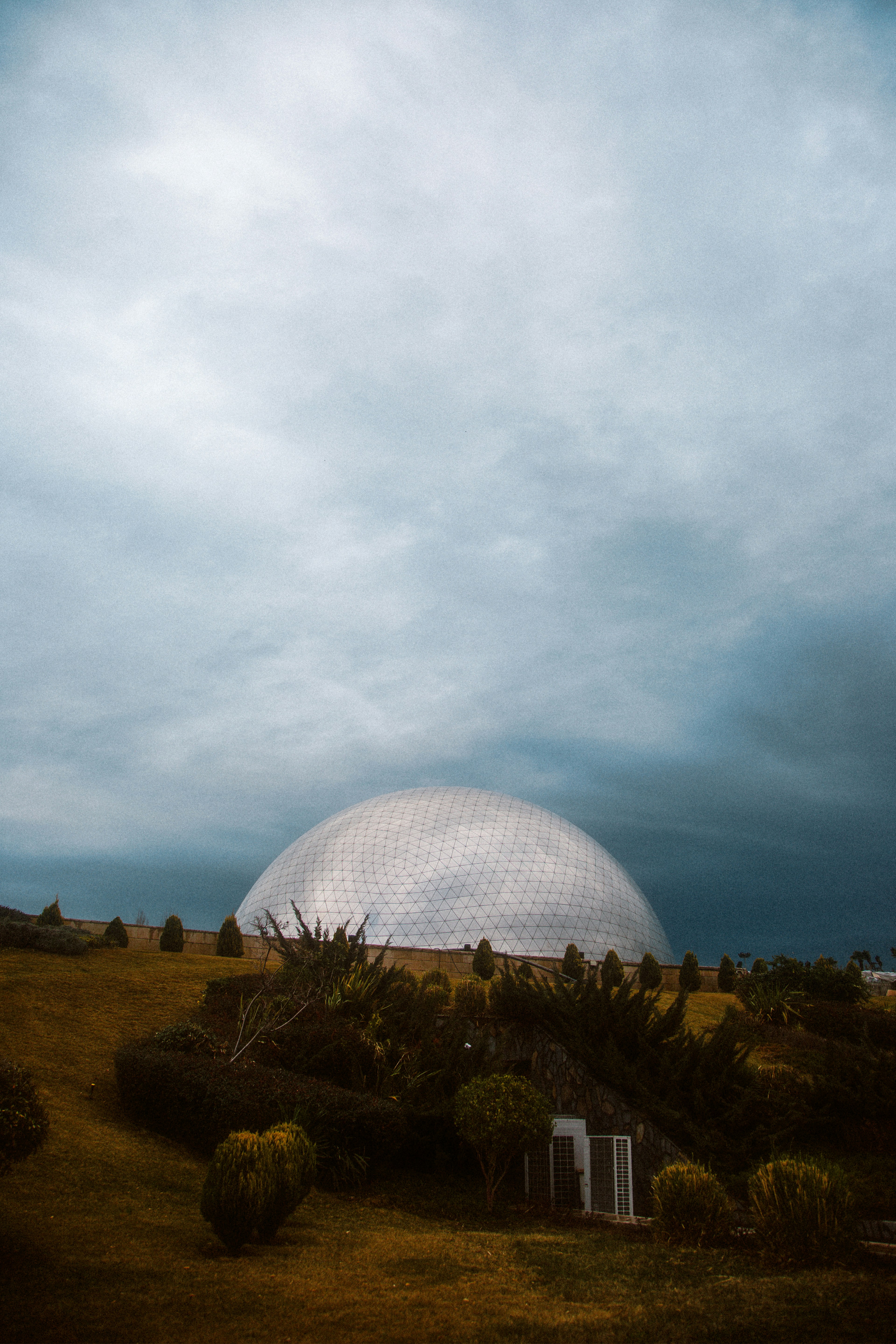 A large, geodesic dome rises against a moody sky, surrounded by lush greenery and subtle landscaping. The scene evokes a sense of mystery and anticipation.
