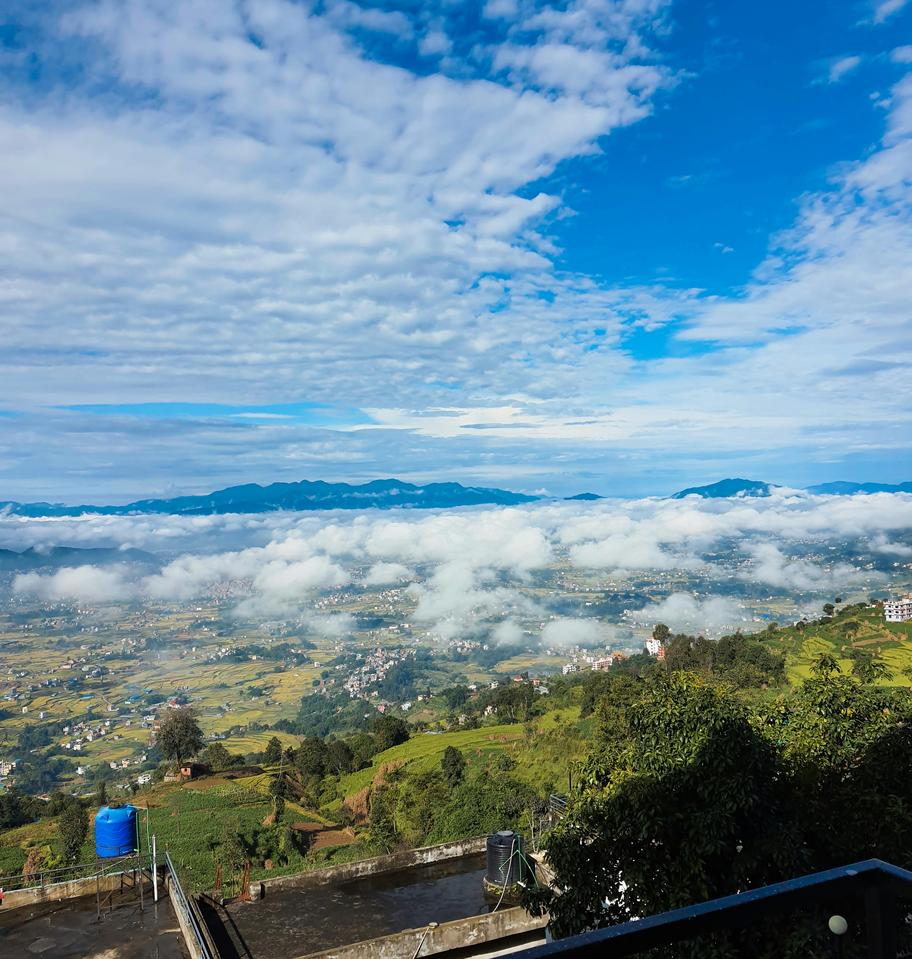 Lush green hills and rolling clouds stretch across the valley under a vibrant blue sky. A water tank and structures hint at human presence amidst the natural beauty.