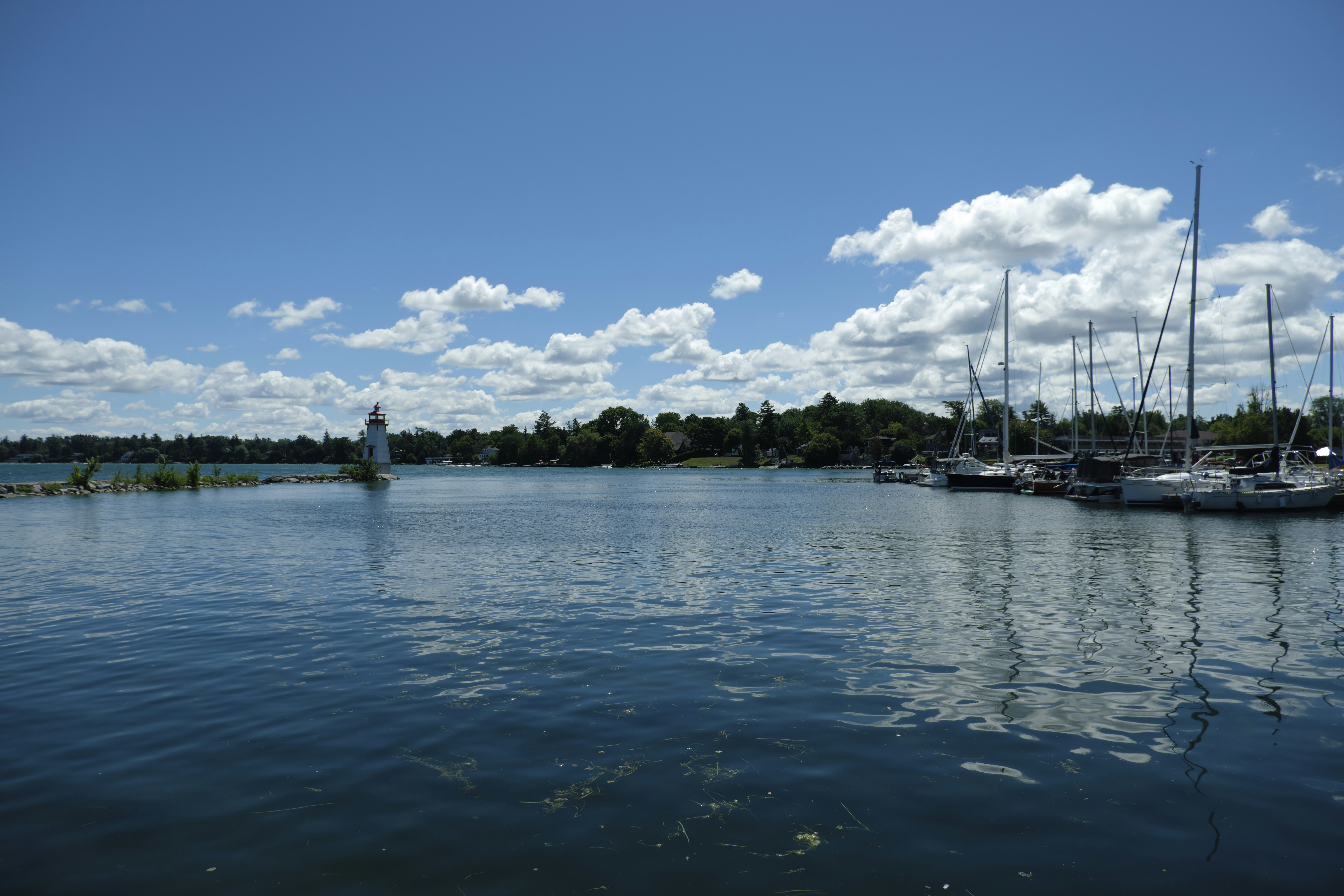 Lighthouse standing tall beside a calm harbor dotted with sailboats under a bright blue sky with fluffy clouds.