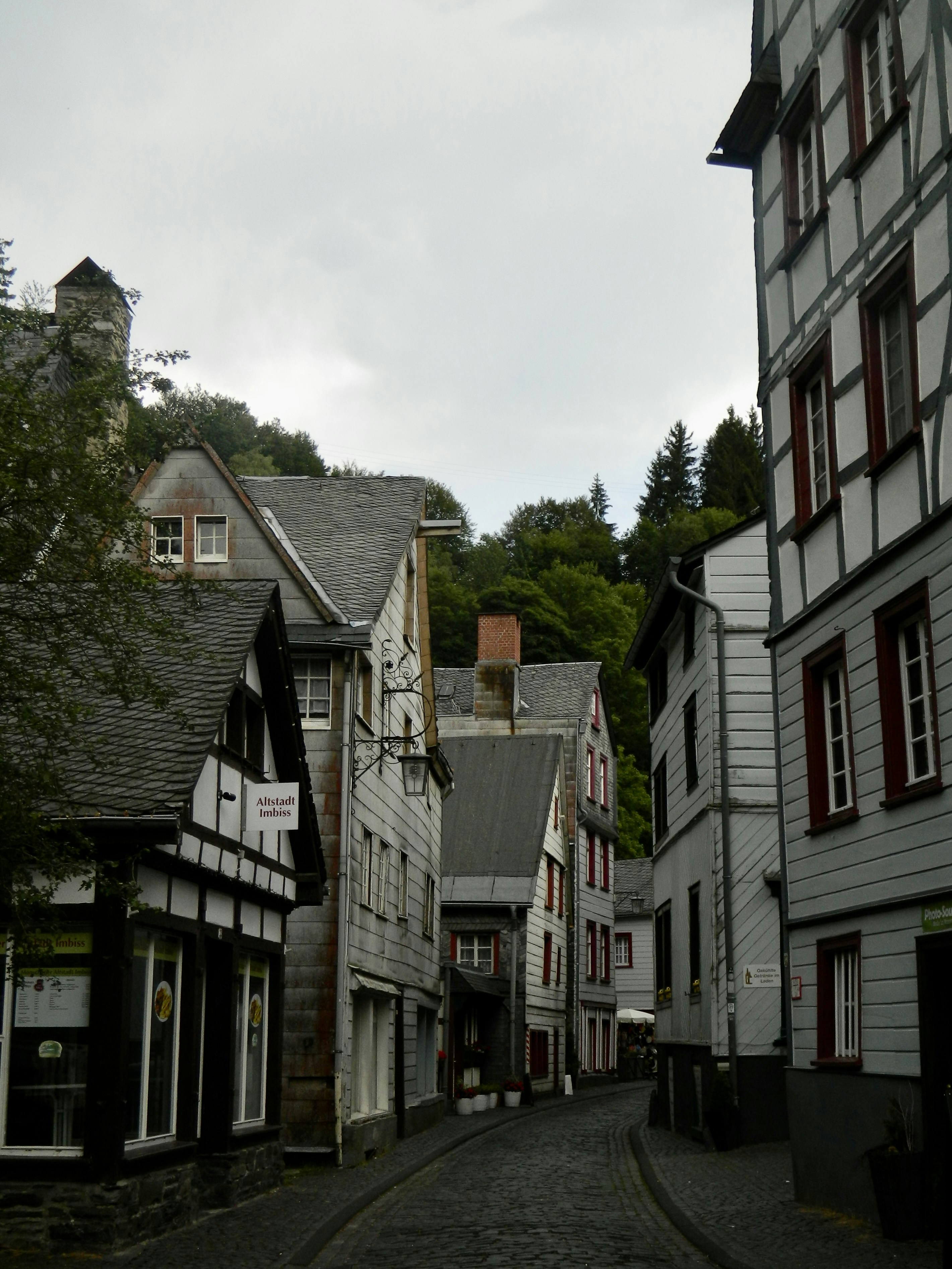 Narrow cobblestone street winds between timber-framed houses with white walls and dark wooden beams under an overcast sky.