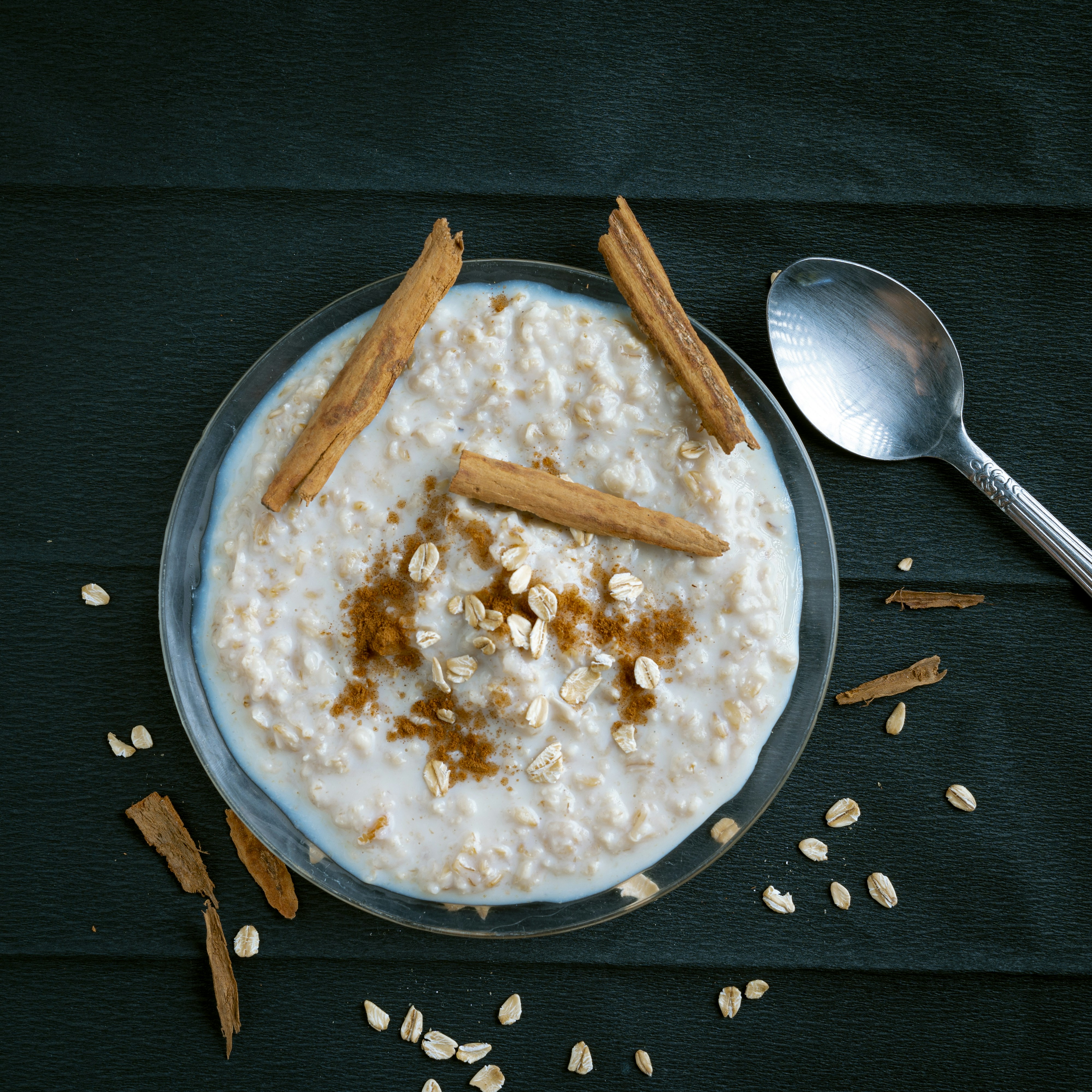 Bowl of creamy oatmeal topped with cinnamon sticks and scattered oats on a dark textured background.