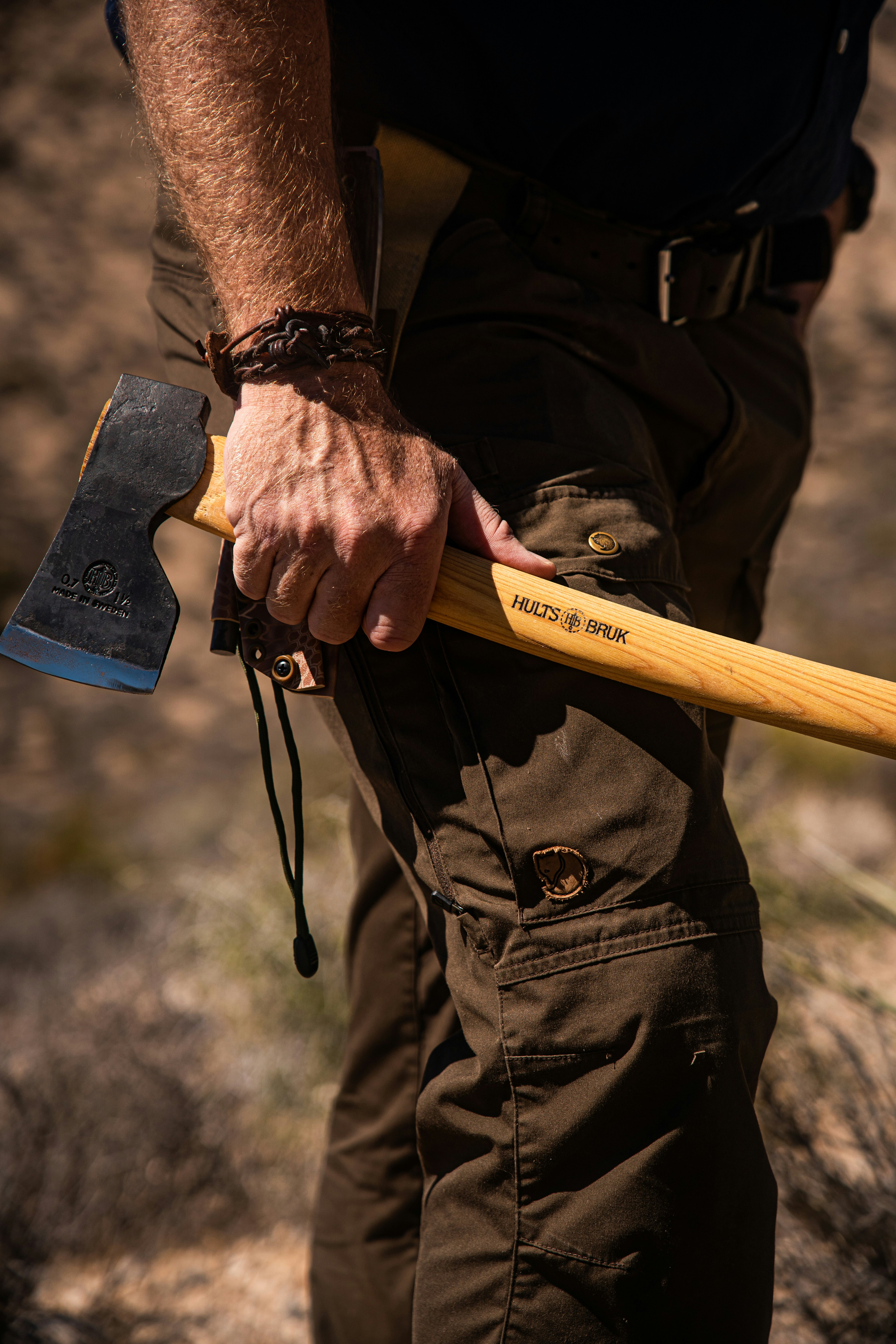 A close-up of a hand gripping a wooden-handled axe, showcasing the rugged texture of the clothing and the tool. The background features natural elements, emphasizing a connection to craftsmanship and outdoor skills.