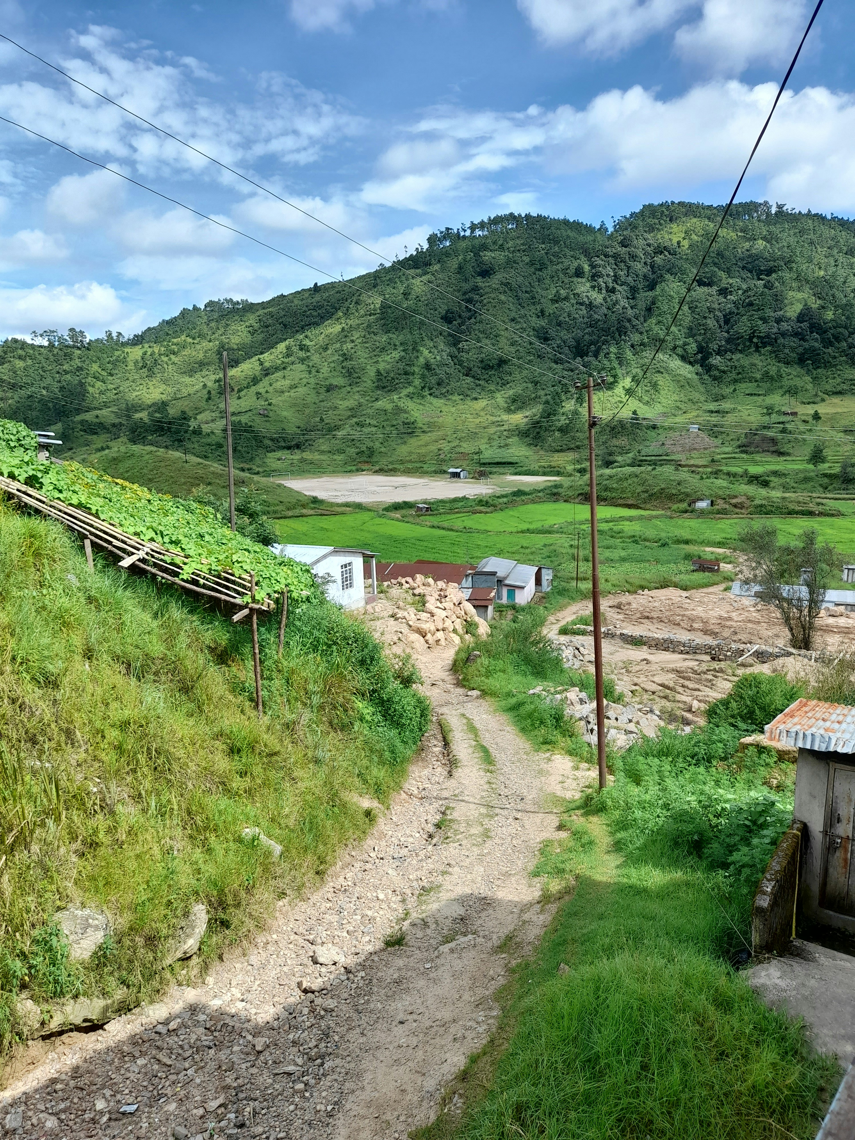 A dirt road snakes up a grassy slope toward terraced fields and modest buildings, framed by green hills. A bright blue sky with scattered clouds completes the rural panorama.