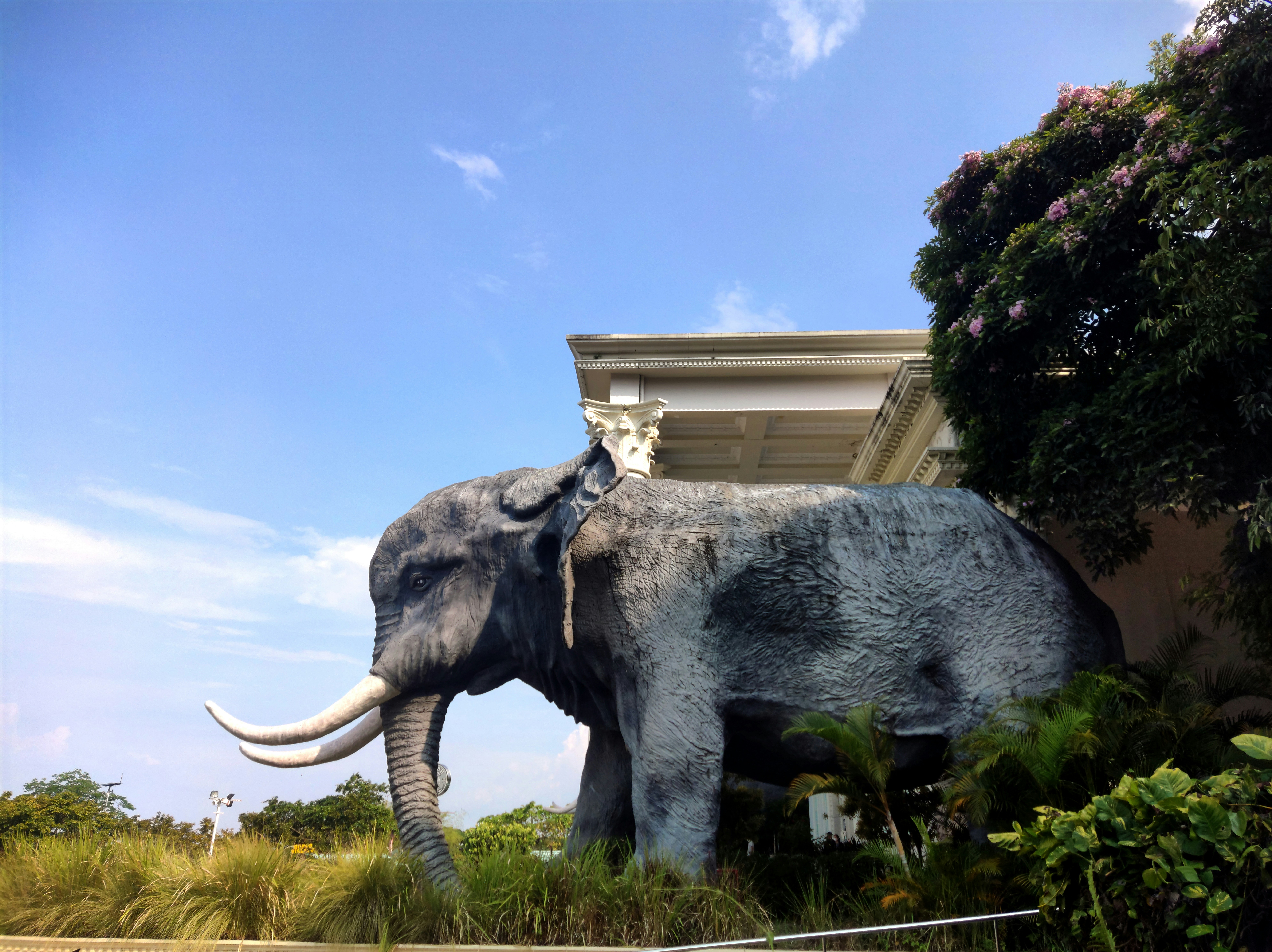A colossal elephant sculpture stands proudly against a clear blue sky, surrounded by lush greenery and architectural details.