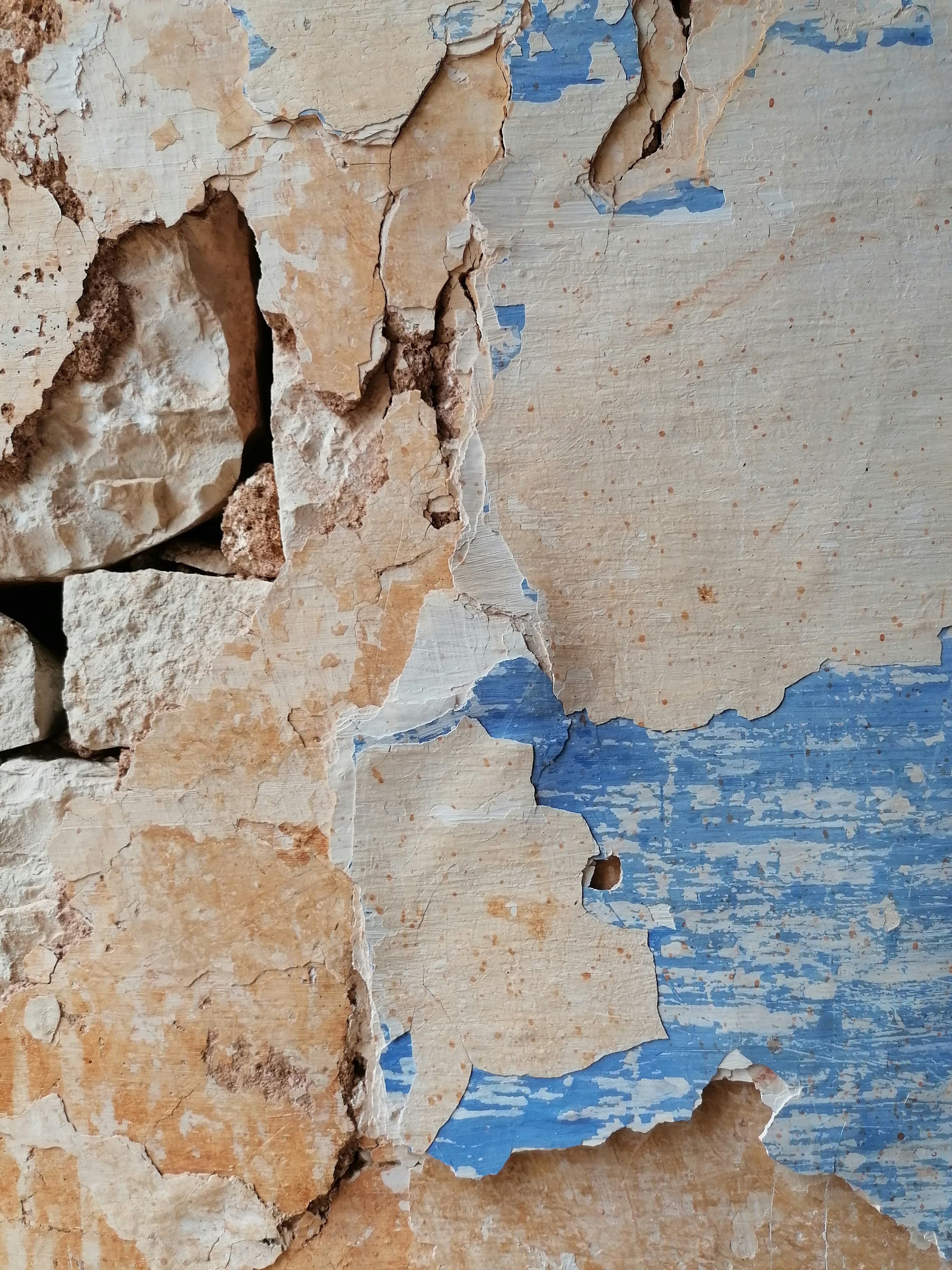 Close-up of a weathered wall where beige plaster peels away to reveal chipped blue paint, creating a layered, textured surface.