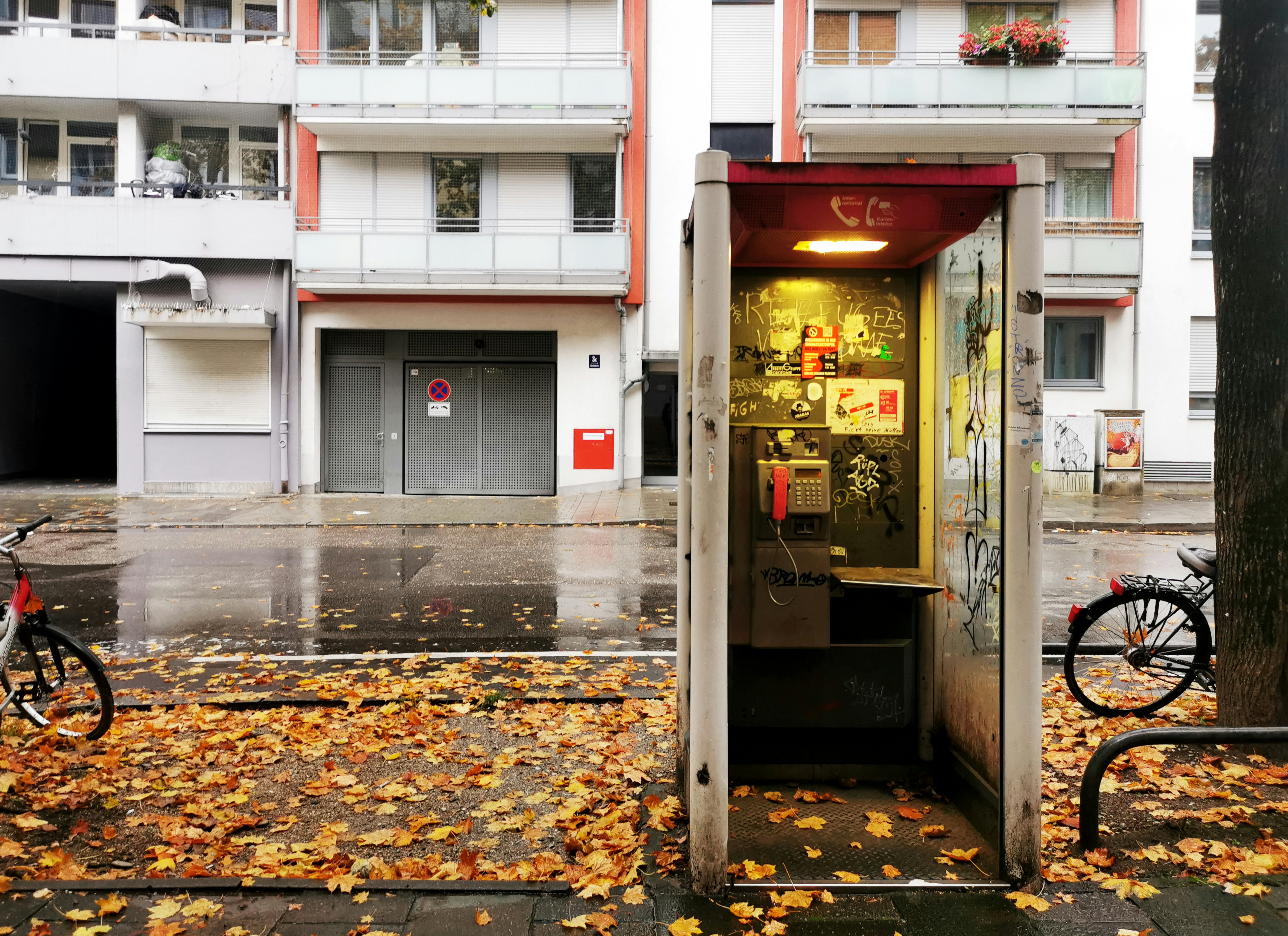 Abandoned telephone booth illuminated by a warm light, surrounded by autumn leaves and urban architecture. Graffiti adorns the booth's interior, hinting at stories untold.