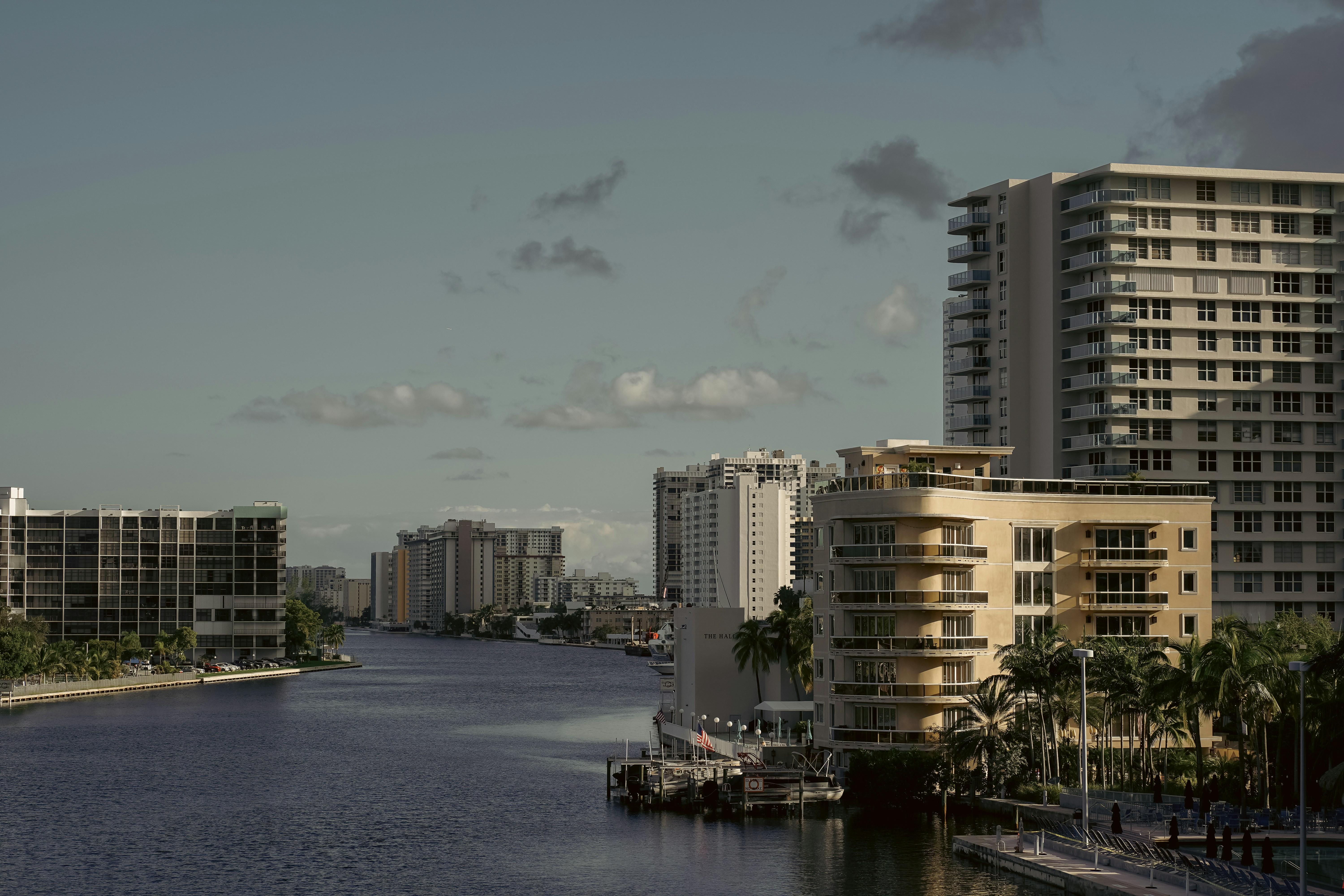 High-rise buildings line a tranquil river under a partly cloudy sky.