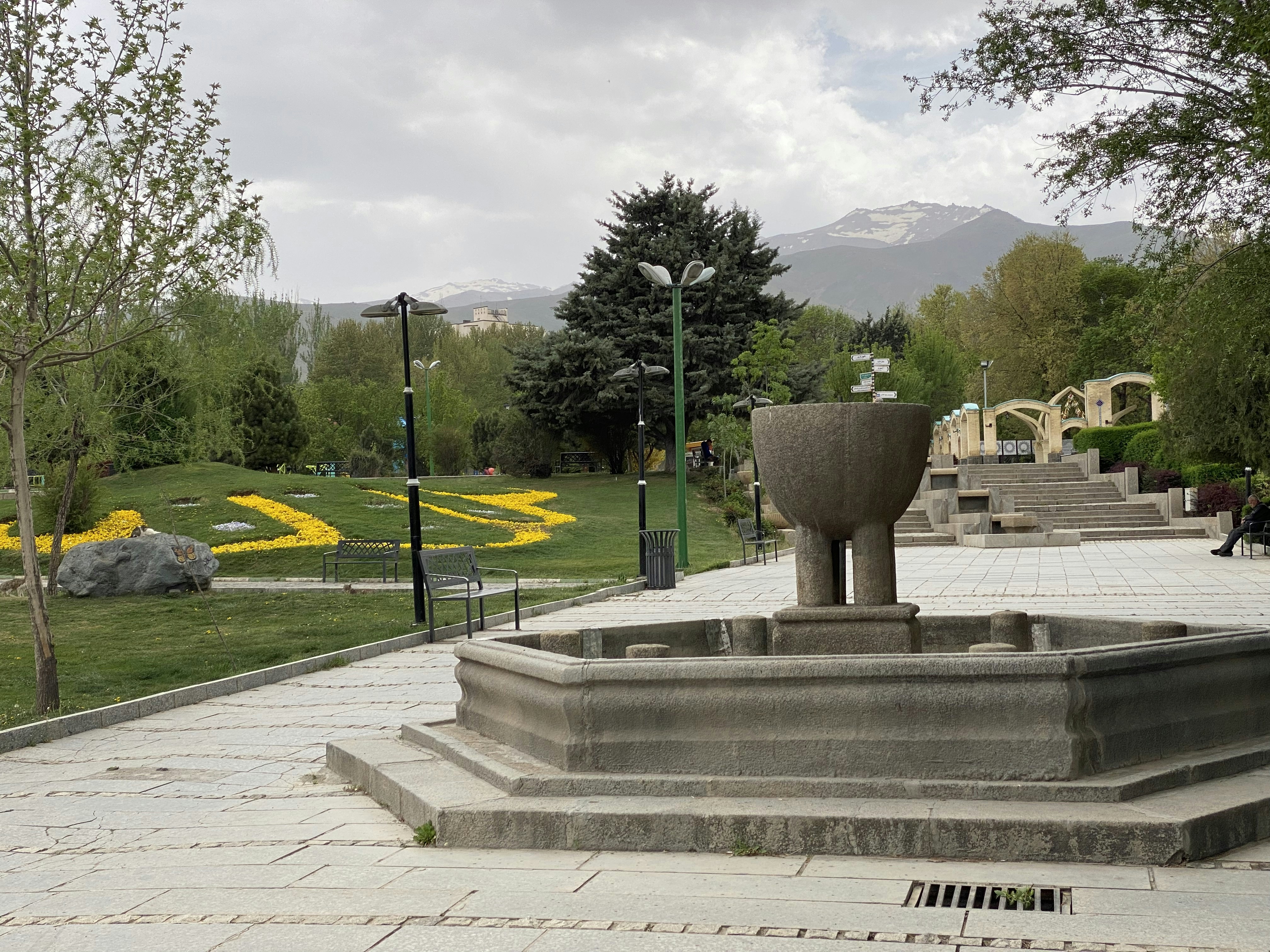 Stone statue in a park with lush greenery and distant mountains under a cloudy sky.