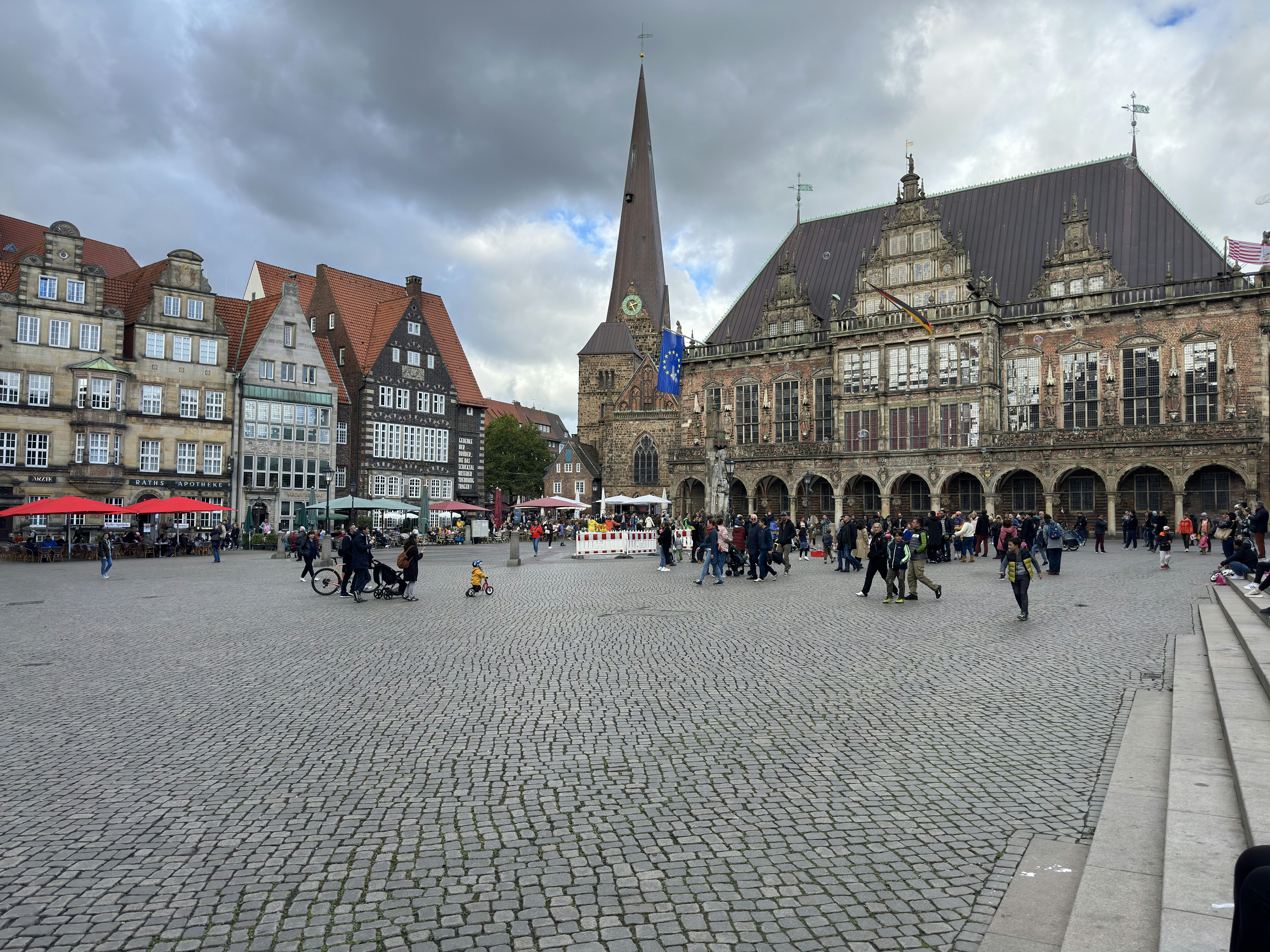 Vibrant town square in Bremen, showcasing historic architecture and bustling crowds. The iconic spire of St. Peter's Church rises prominently against a cloudy sky.
