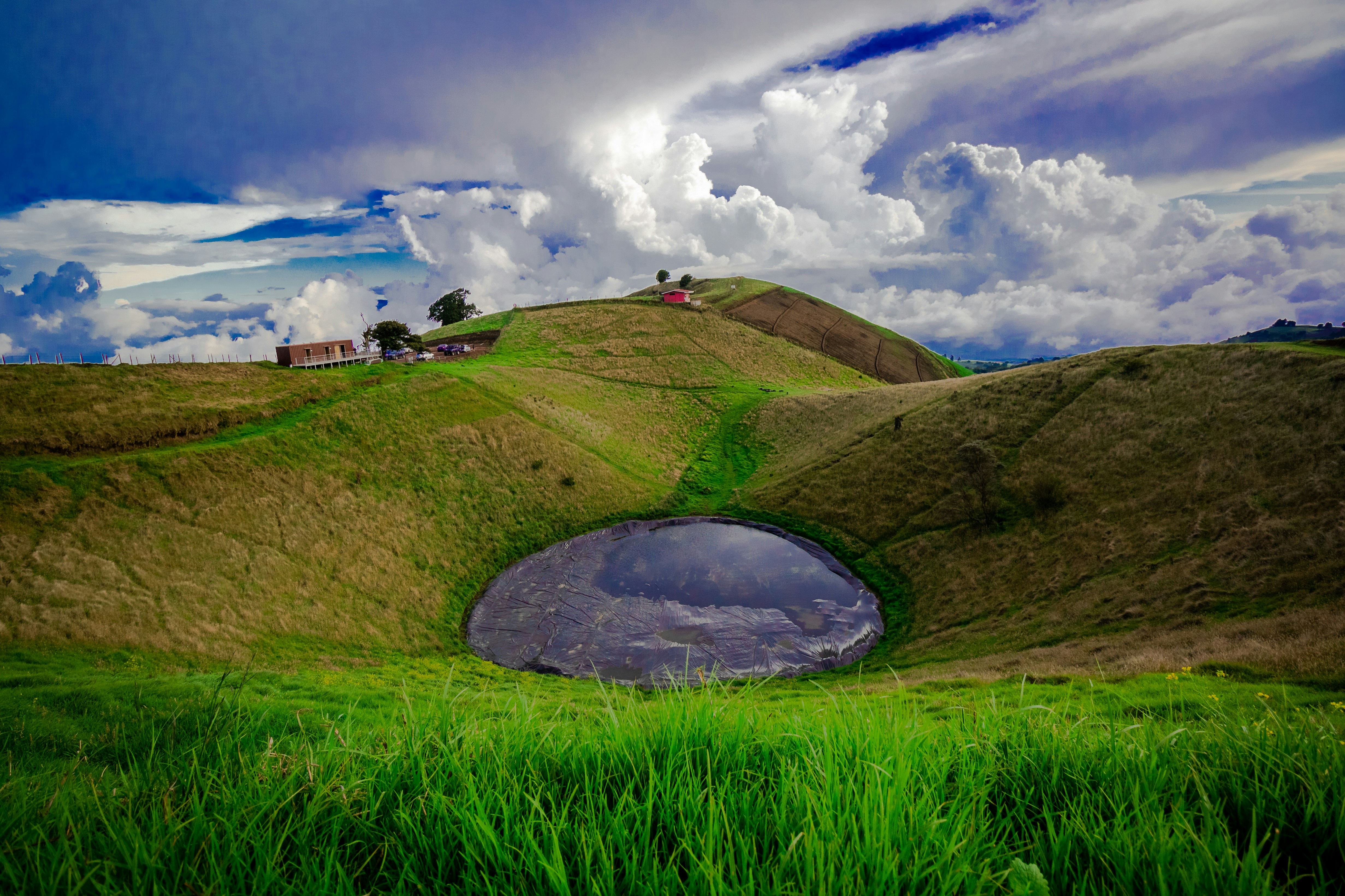 Grassy crater with a reflective pond surrounded by rolling hills under dramatic clouds.