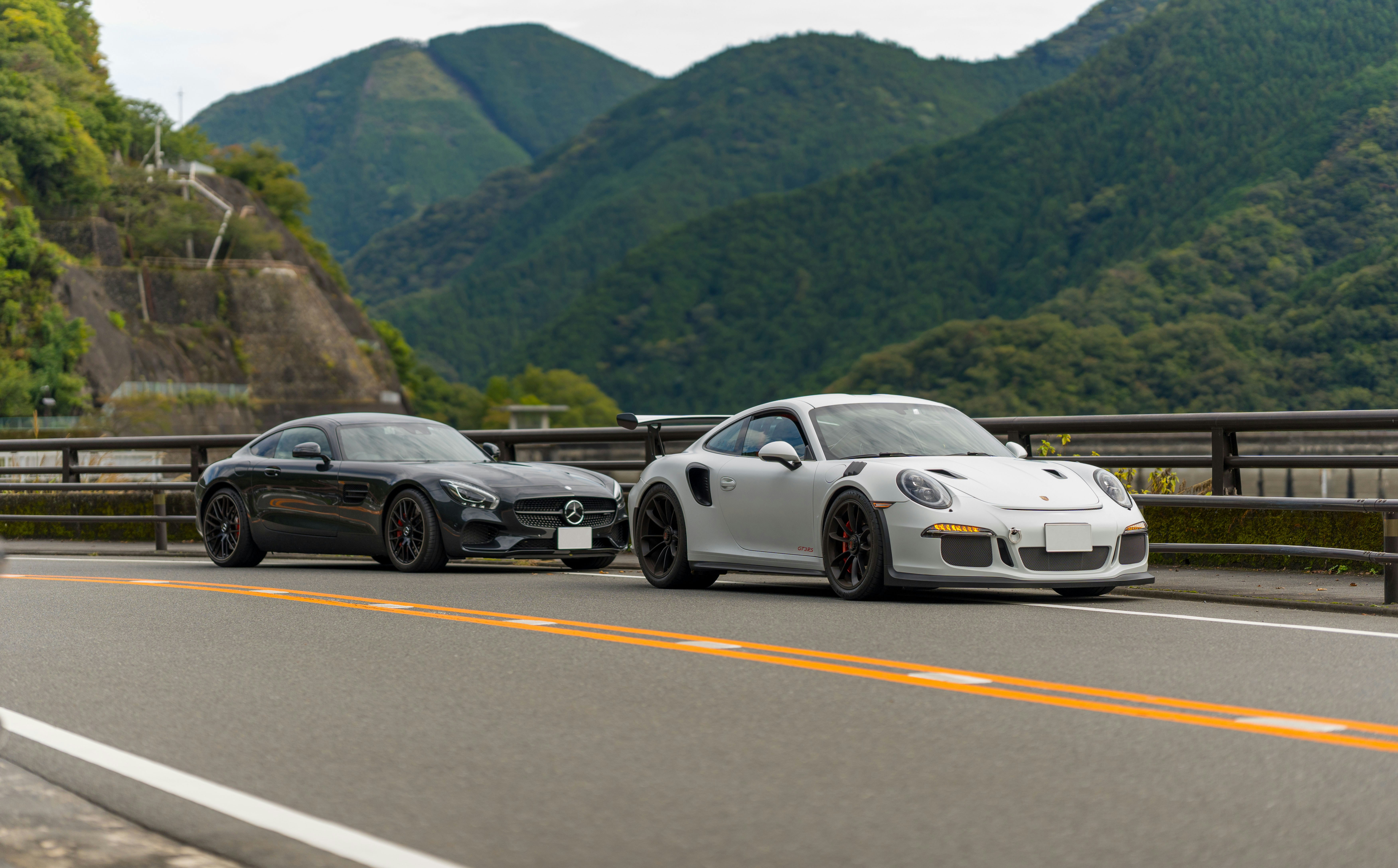 A sleek black Mercedes-Benz and a striking white Porsche 911 parked on a scenic road with lush green mountains in the background.
