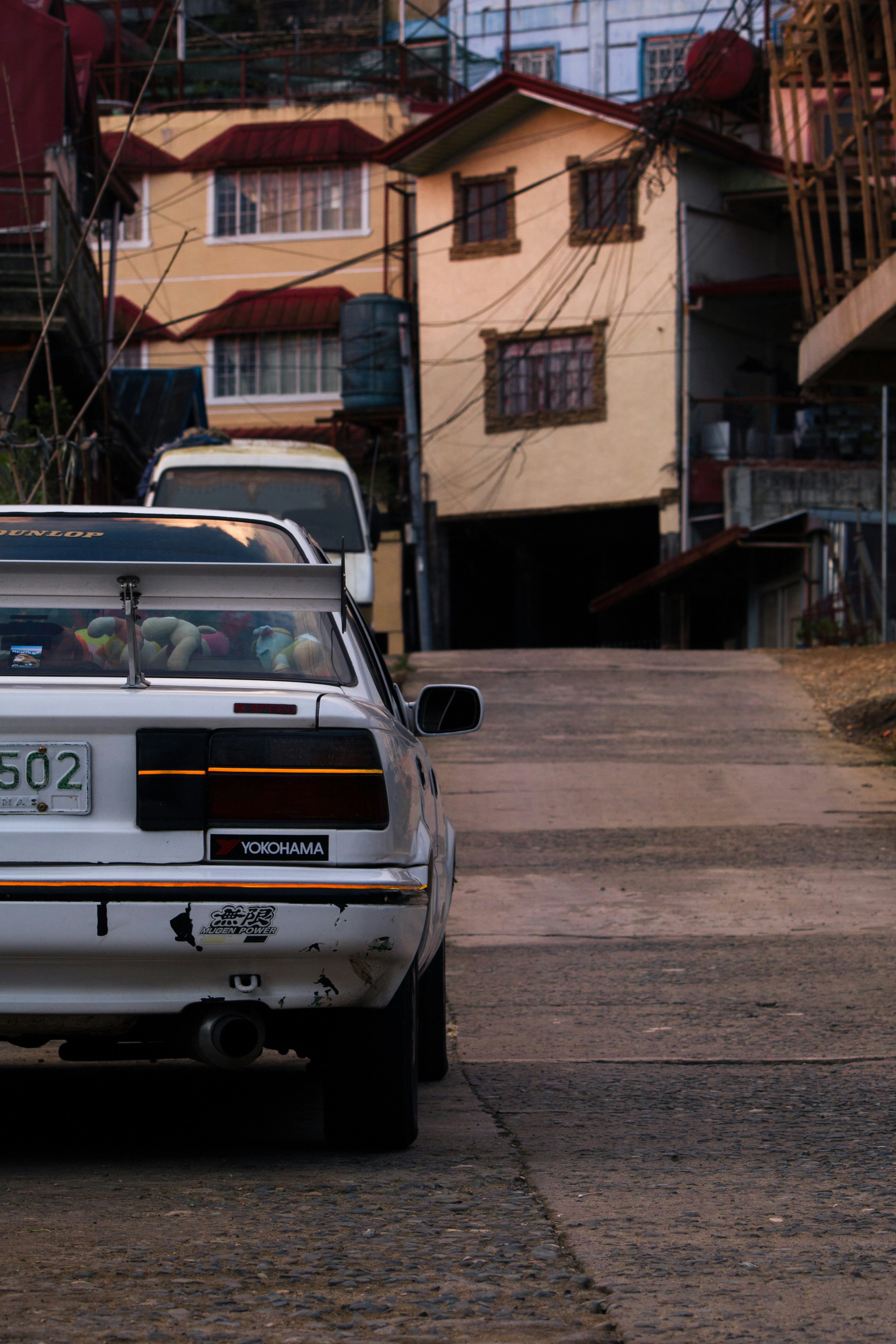 Rear-Quarter View of a White Sedan on a Cobblestone Street, Framed by Colorful Barrio Houses