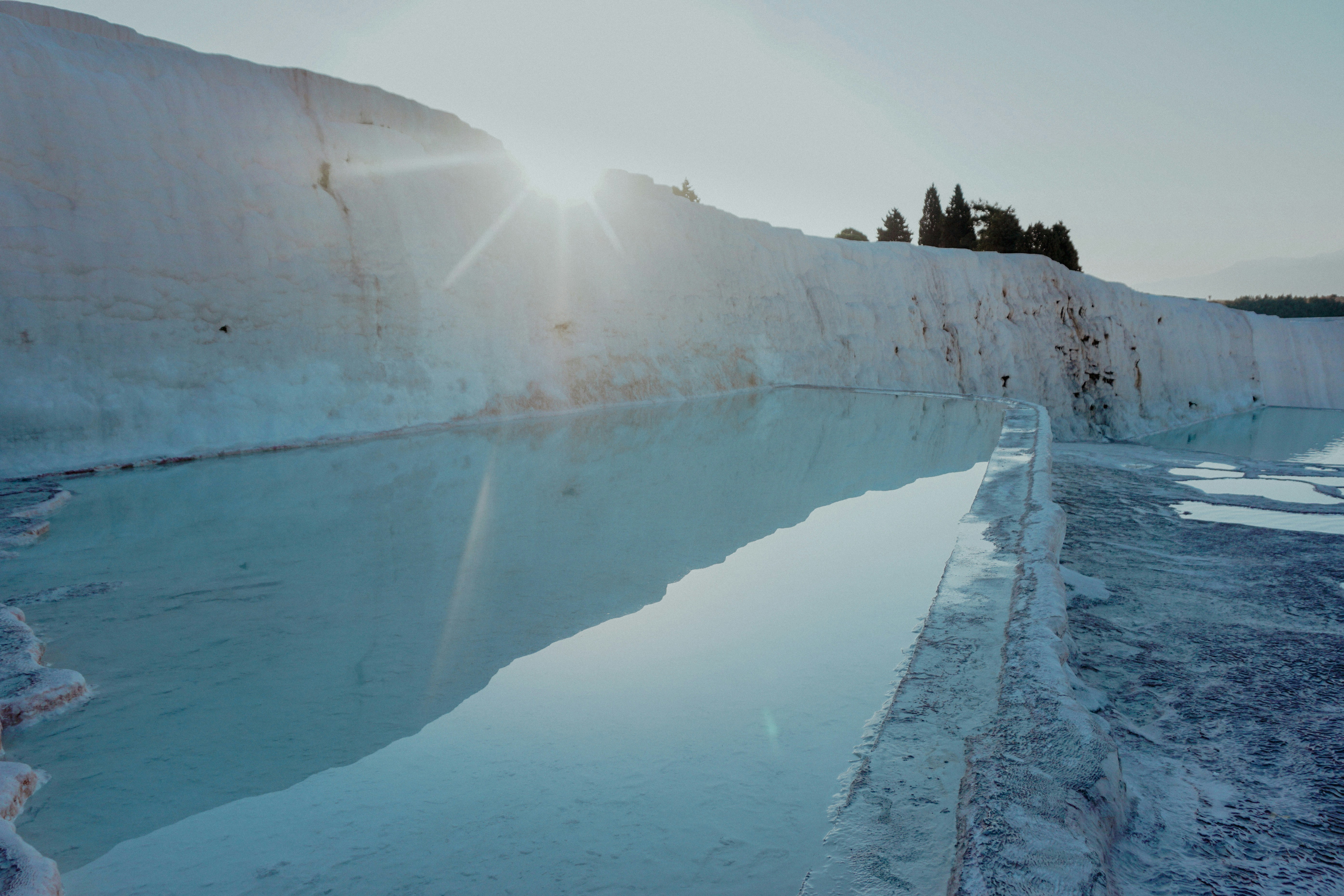 Calm water reflecting sunlight beside a rocky ledge with distant trees under a clear sky.
