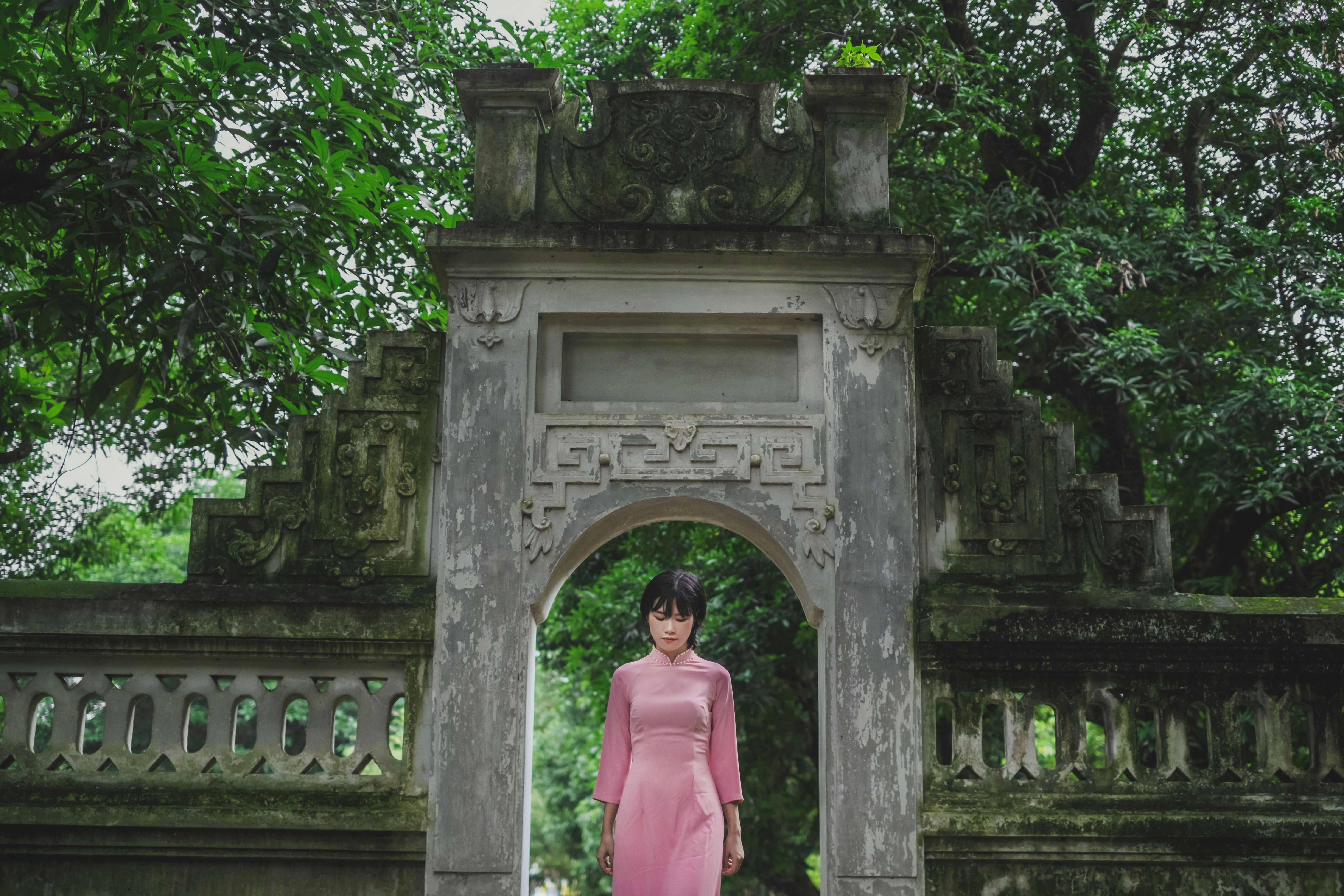 A young woman in a pink dress stands gracefully beneath an ornate stone archway, surrounded by lush greenery.