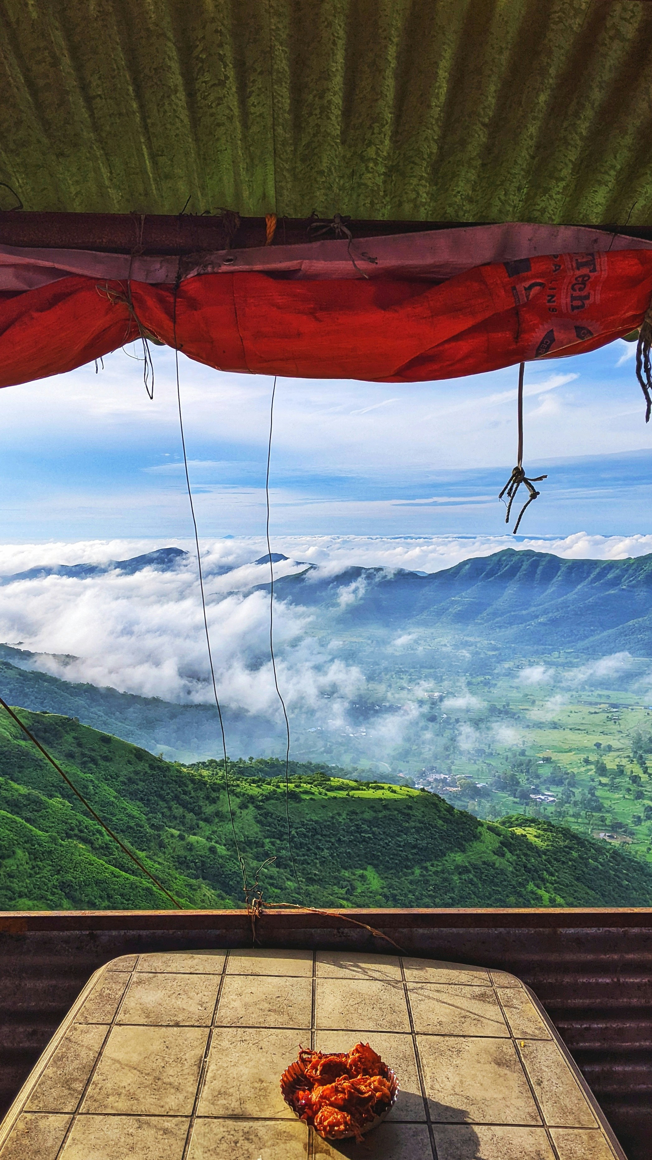 View from a rustic shelter overlooking lush green mountains and clouds, with a colorful offering placed on a table in the foreground.