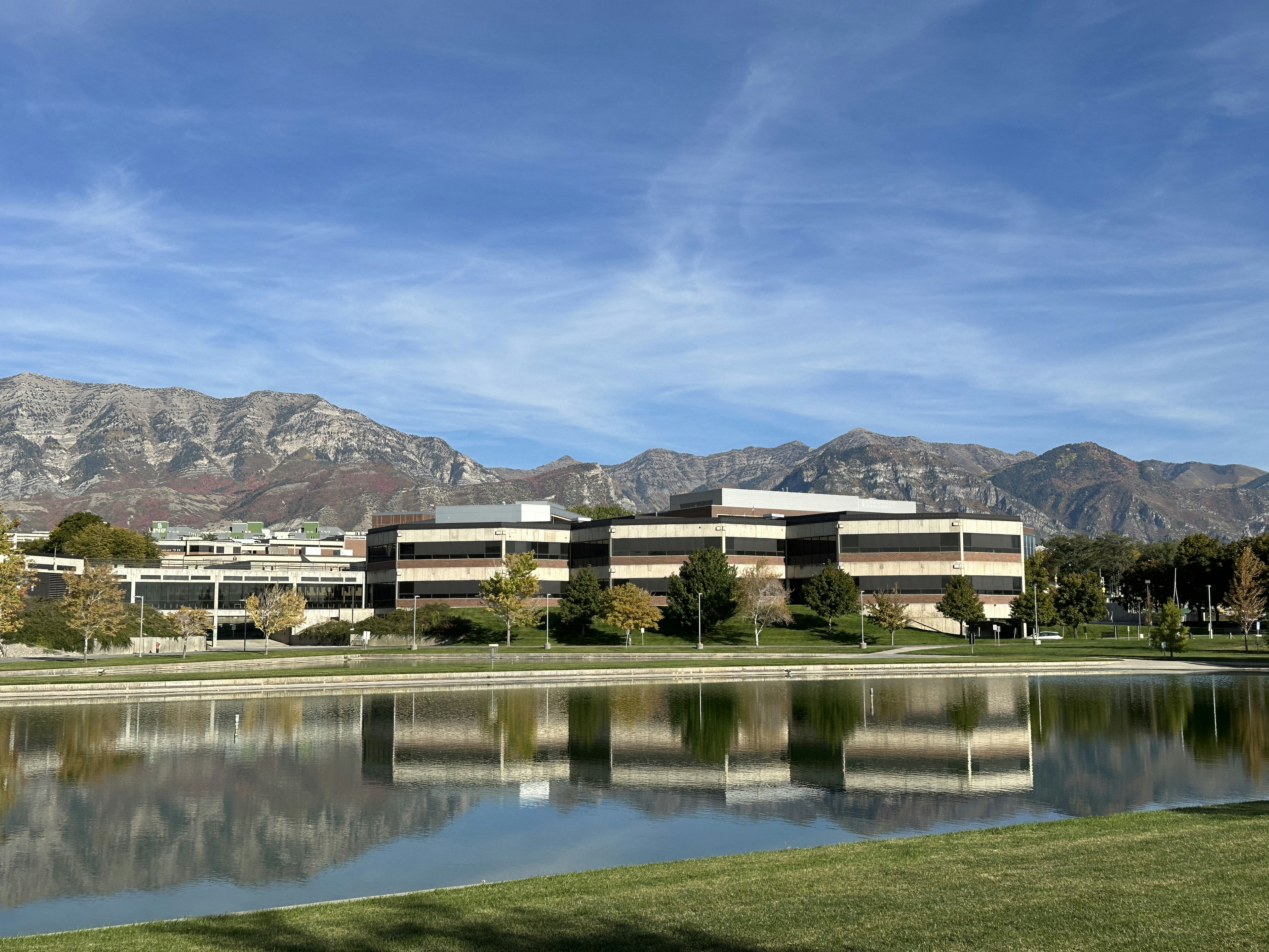 Modern campus buildings mirrored in a tranquil pond with mountain backdrop under a clear blue sky.