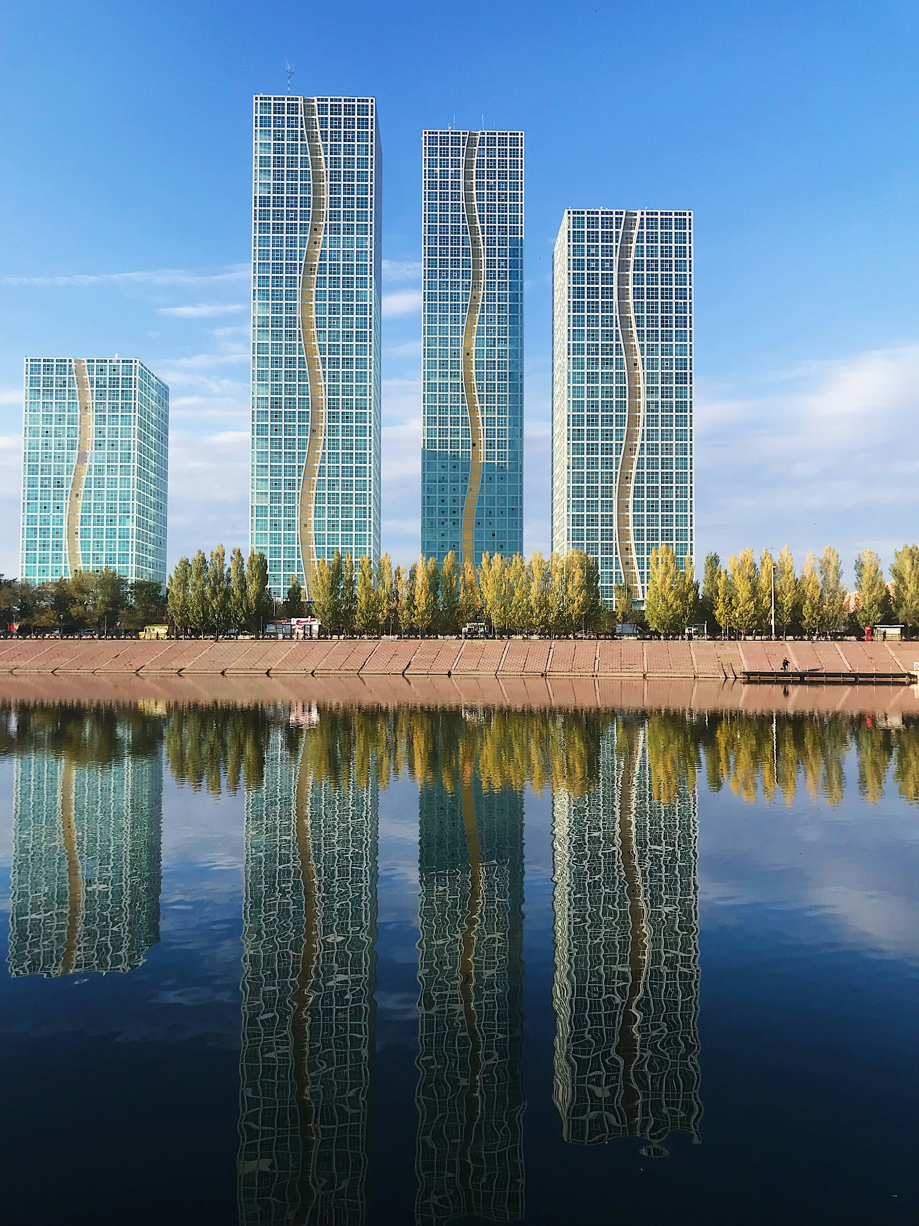 Four modern skyscrapers with unique wavy designs reflect in a calm body of water, framed by golden autumn trees along the shore.
