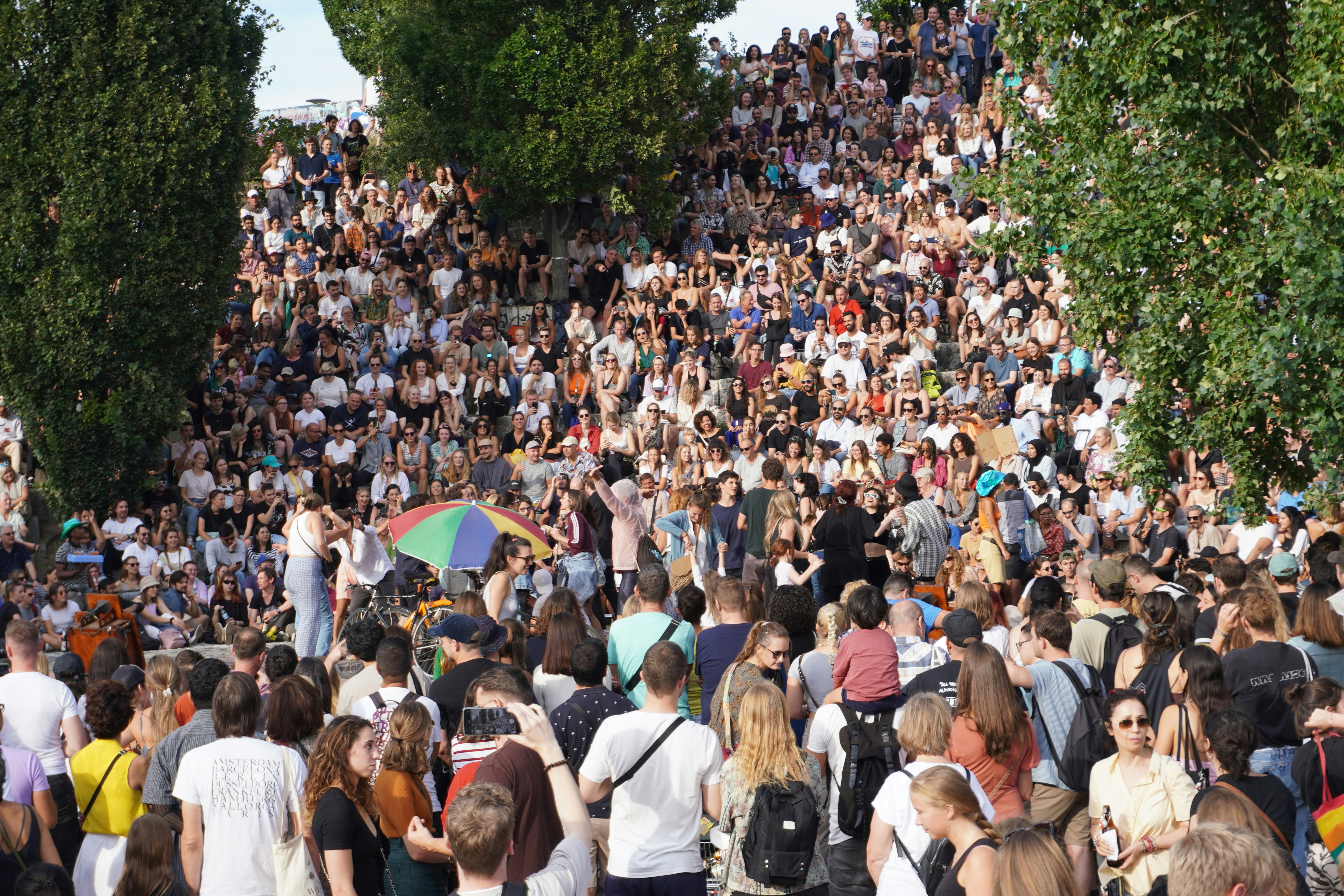 Crowd of diverse individuals gathered on a hillside, enjoying a lively event under the sun.