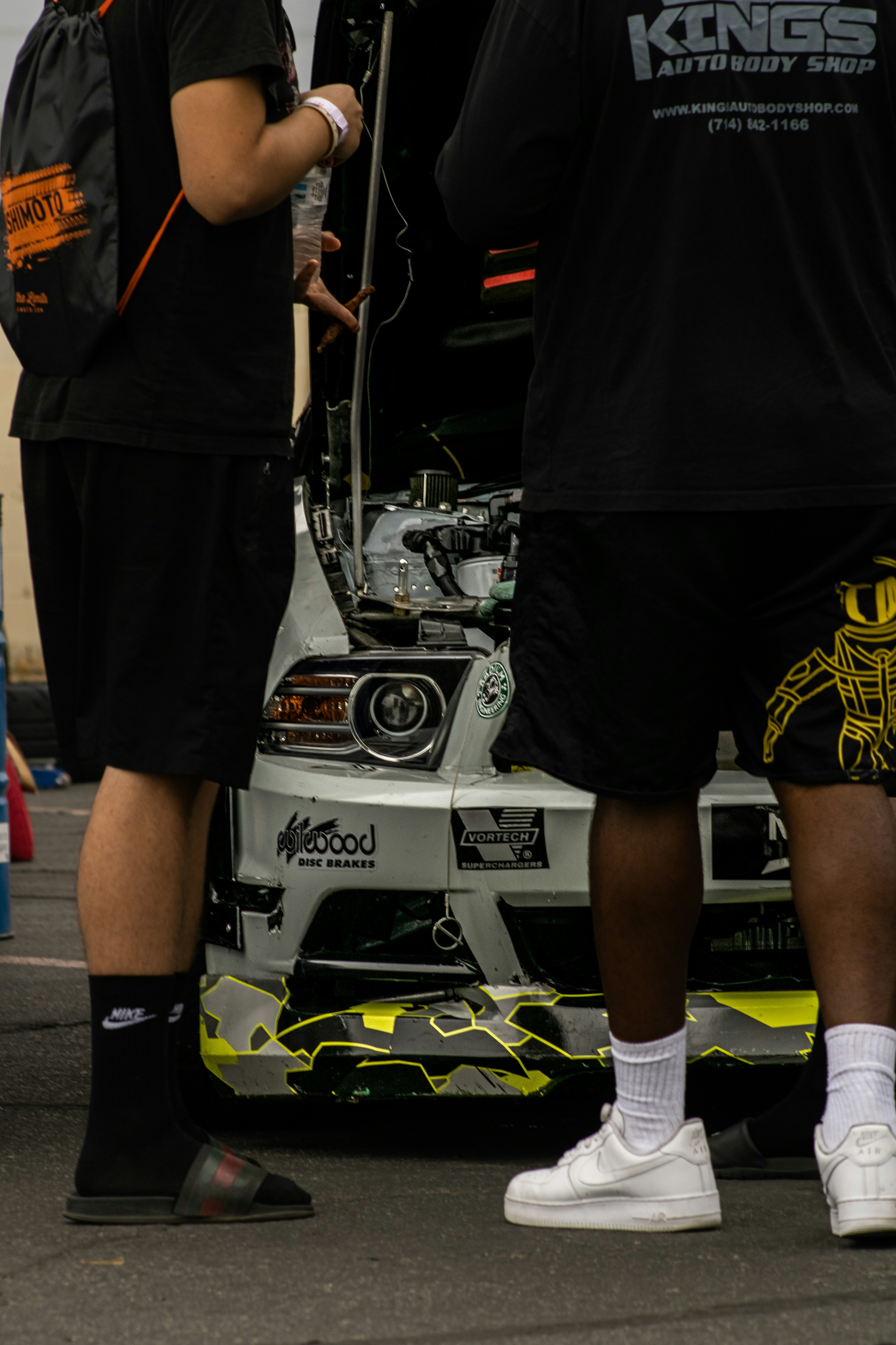 Dramatic scene of crew members gathered around a modified race car with its hood open, exposing the engine bay and front end details.