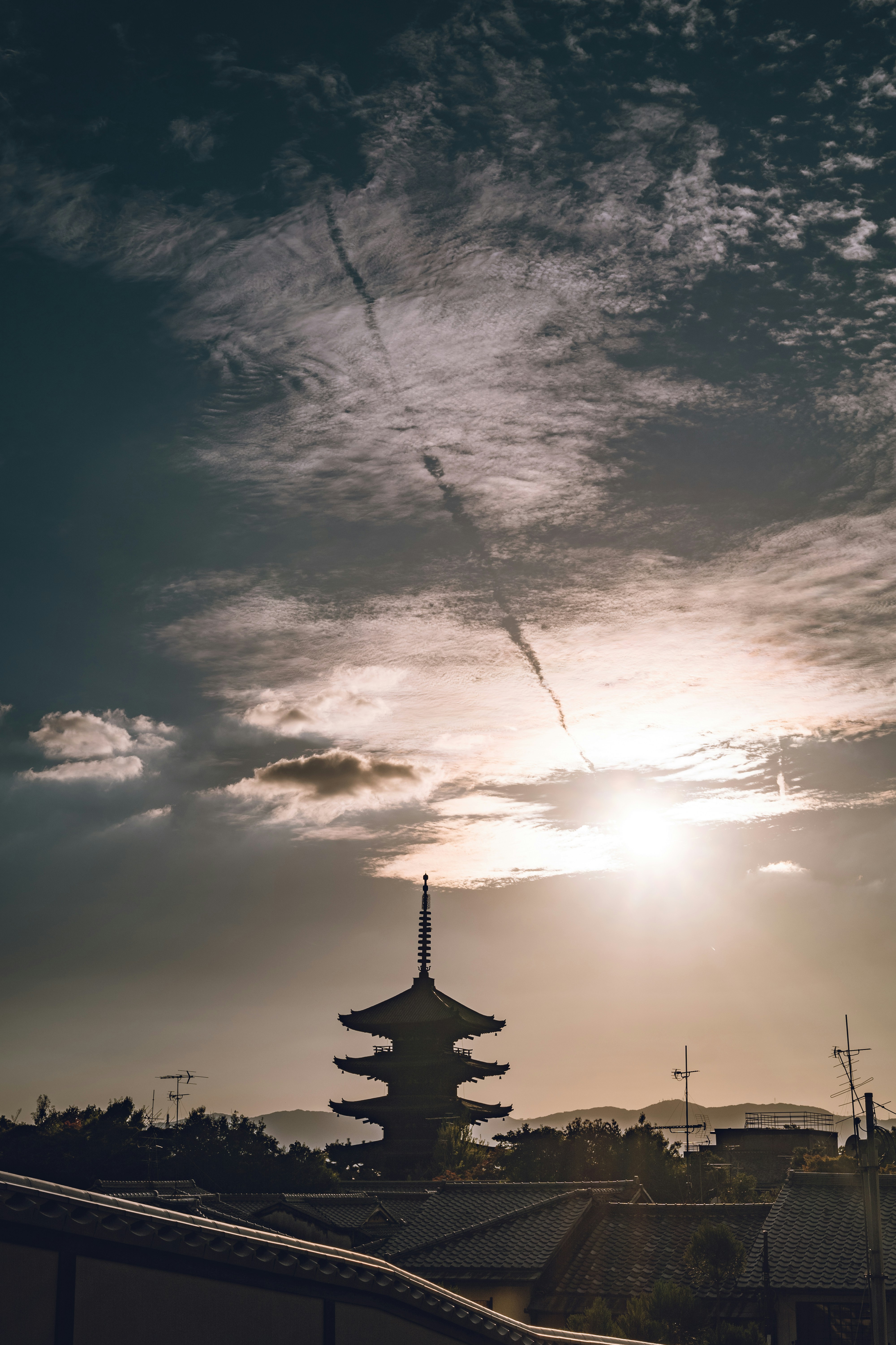 Pagoda silhouette rises above traditional rooftops at sunset, set against a dramatic sky. The scene emphasizes architectural form and warm backlight from the sun.