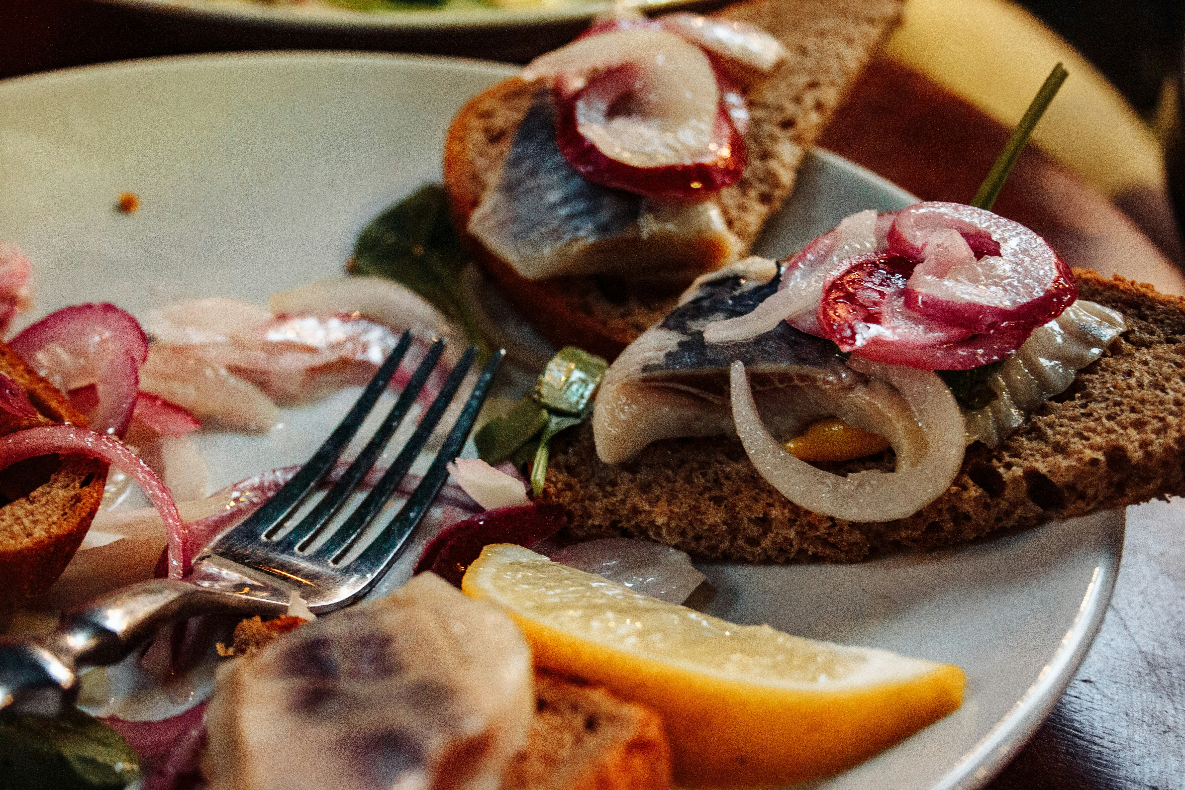Open-faced sandwich featuring herring, pickled onions, and fresh herbs on dark rye bread, accompanied by lemon slices.