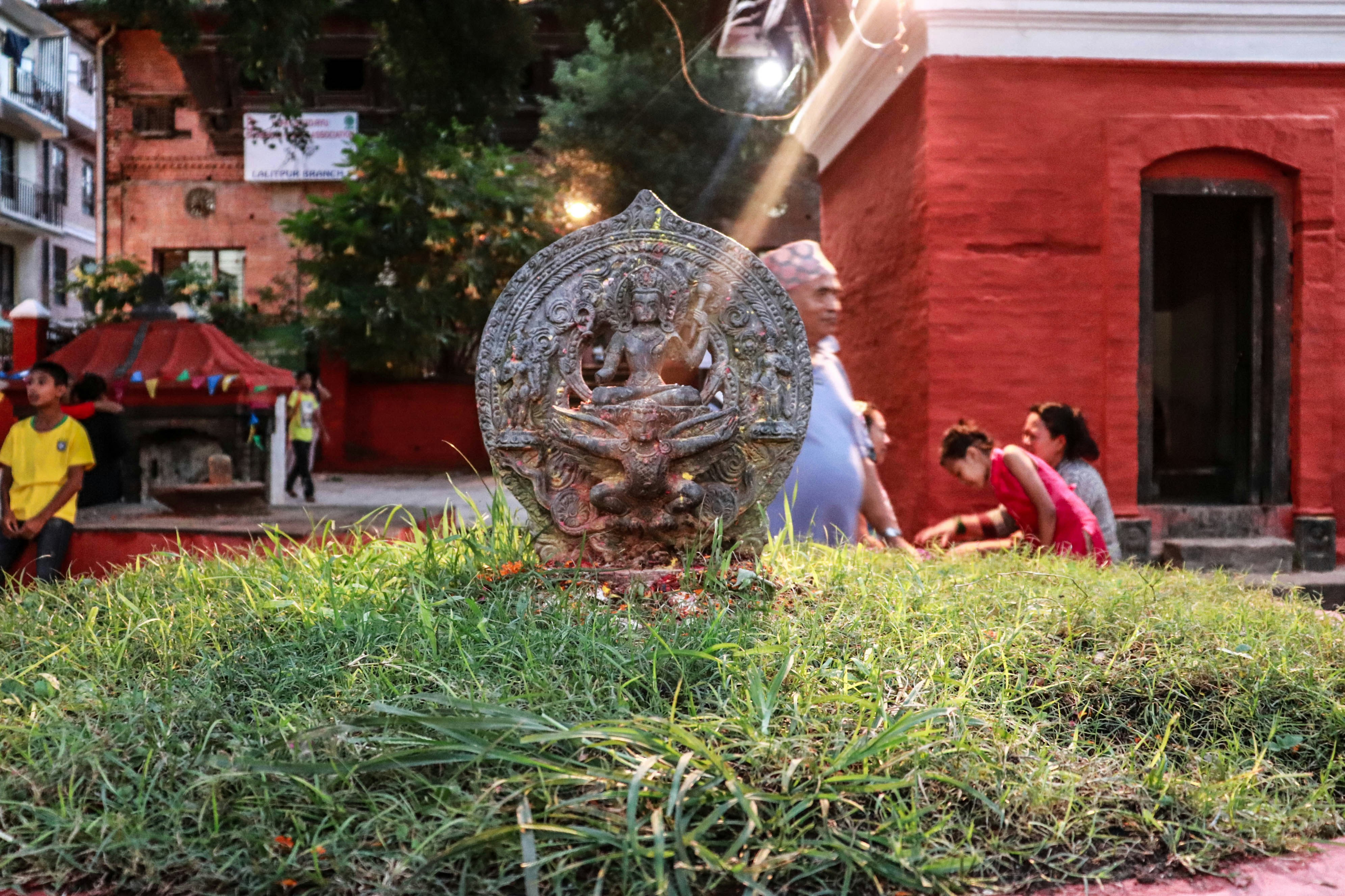 Intricate sculpture surrounded by greenery with people in the background, illuminated by warm evening light.