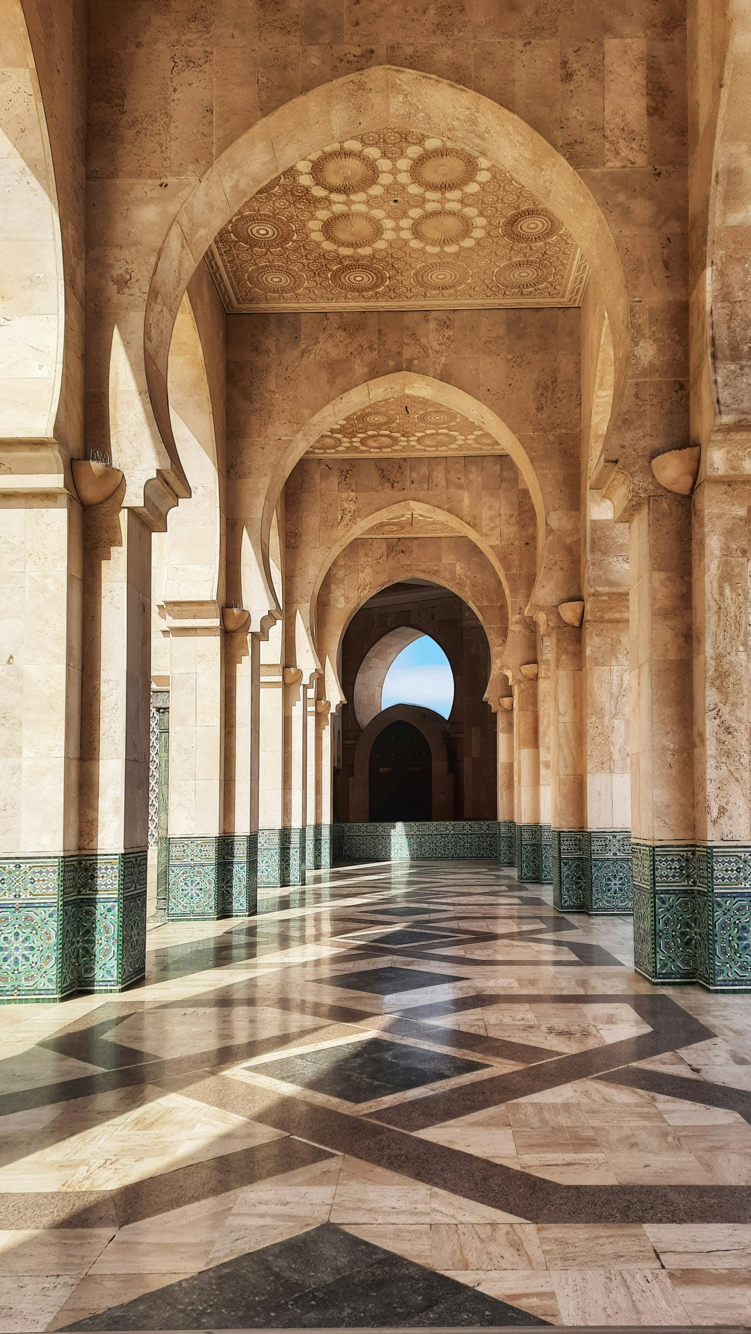 Intricate arches and tiled patterns create a mesmerizing corridor in a grand architectural space. Natural light casts dynamic shadows across the polished floor.